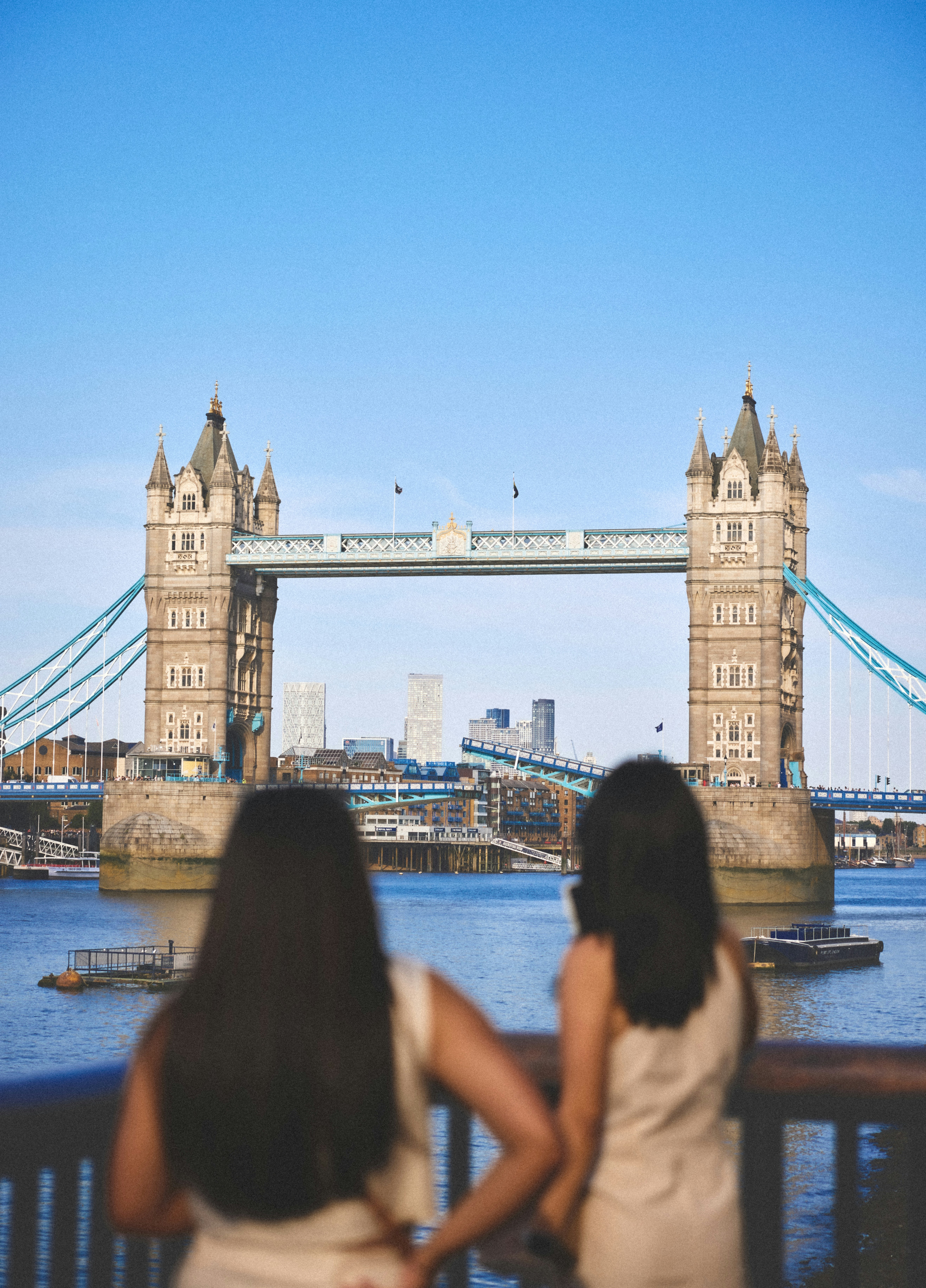 Two women stand at a railing, foregrounding Tower Bridge against the Thames and distant cityscape.