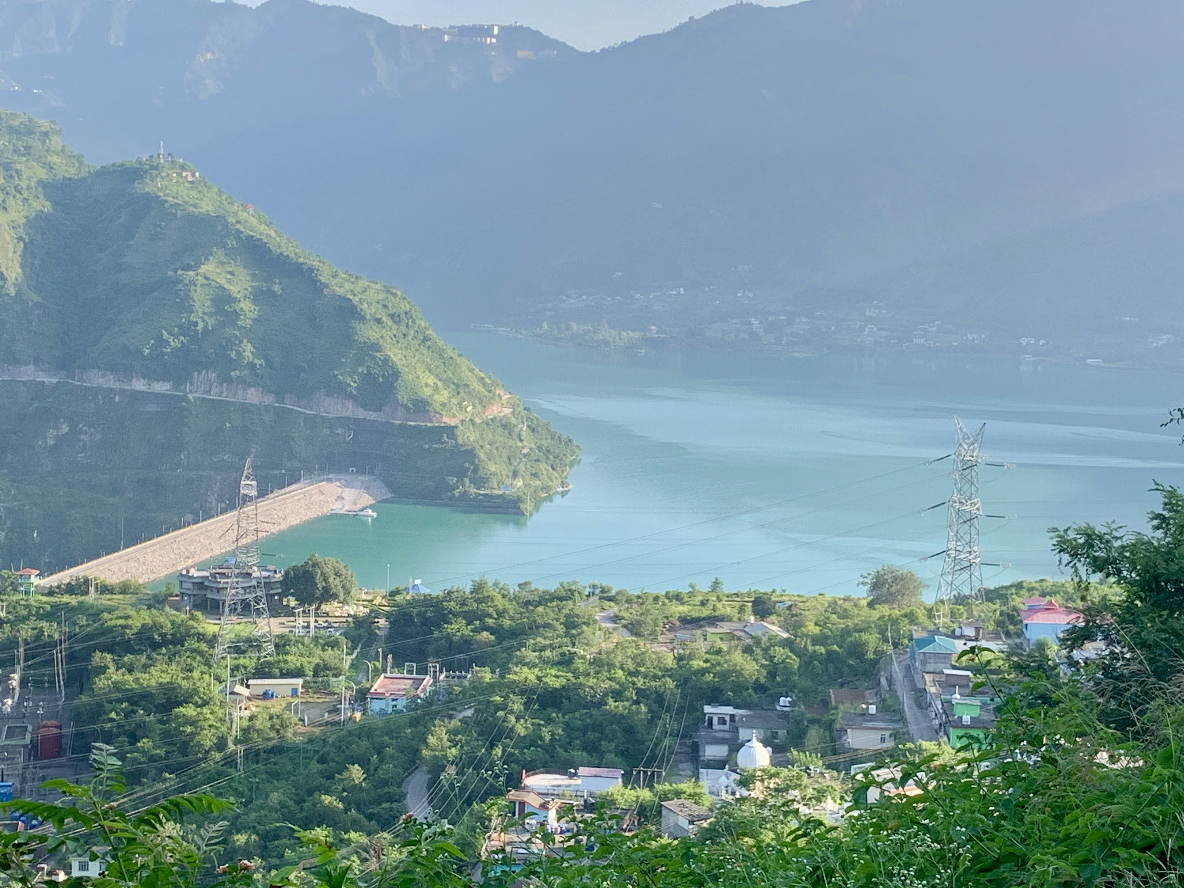 Hillside town with a turquoise reservoir and a curving bridge, seen from an elevated viewpoint; a calm lakeside scene of greenery, rooftops, and distant mountains.