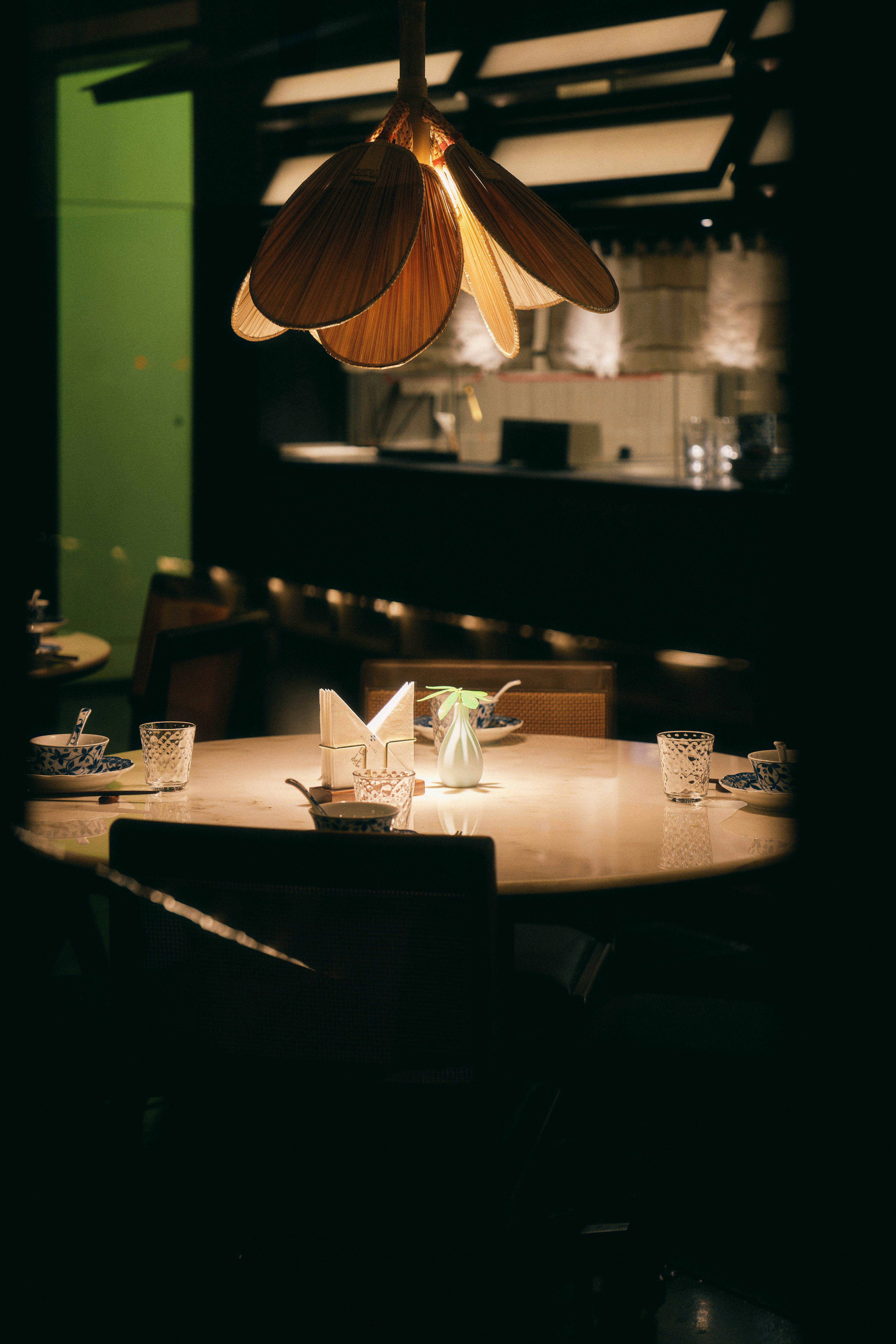 A softly lit dining table under a wooden pendant lamp, with place settings and a small centerpiece visible; the background shows a bar area in a darkened restaurant.