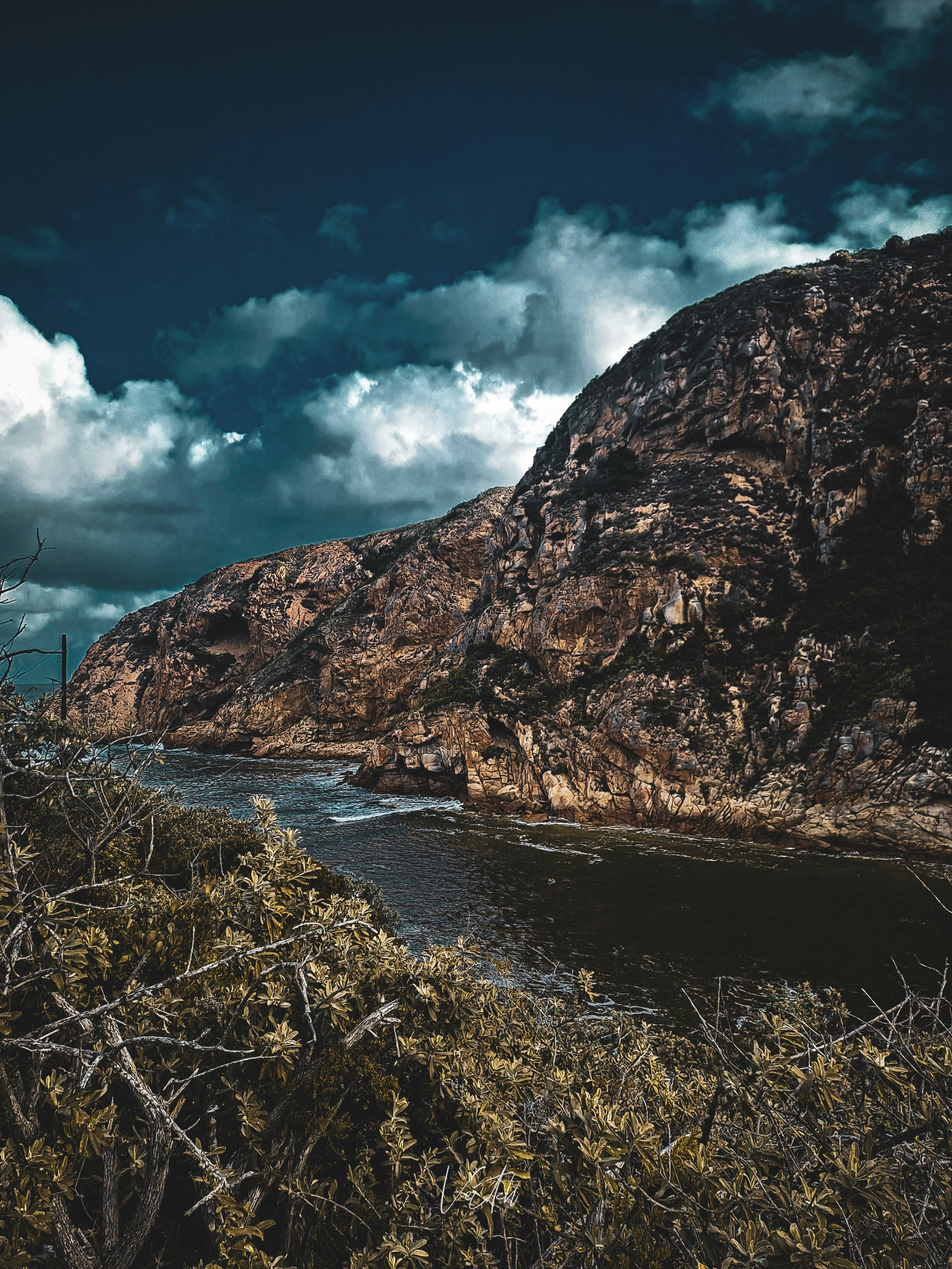 A rocky cliff face overlooking a dark ocean inlet under a cloudy sky