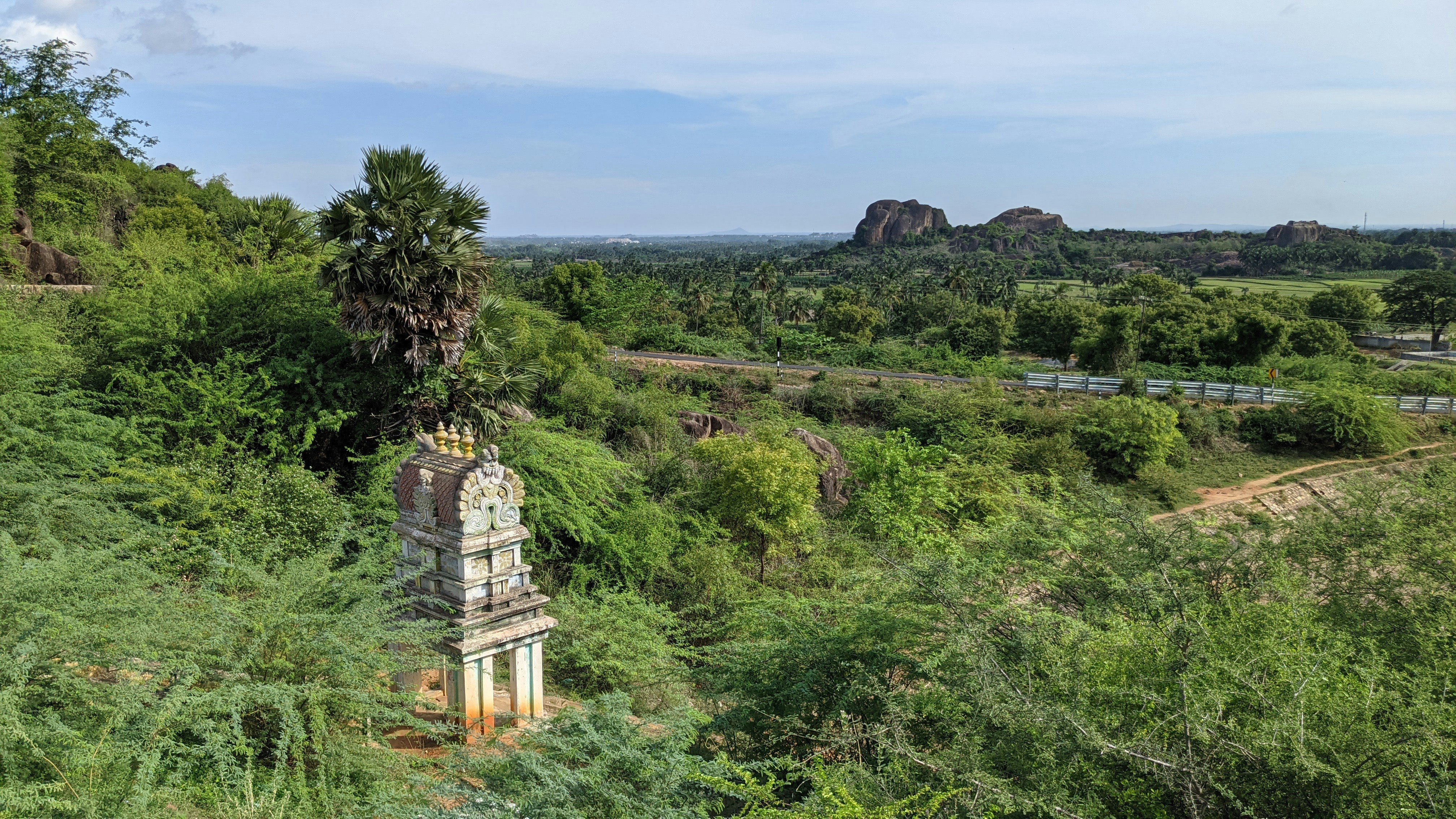 Stone reliquary tower nestled among dense green shrubbery with distant rock formations and a winding road in the background. A serene, landscape scene highlighting historical relics within a lush, rural setting.