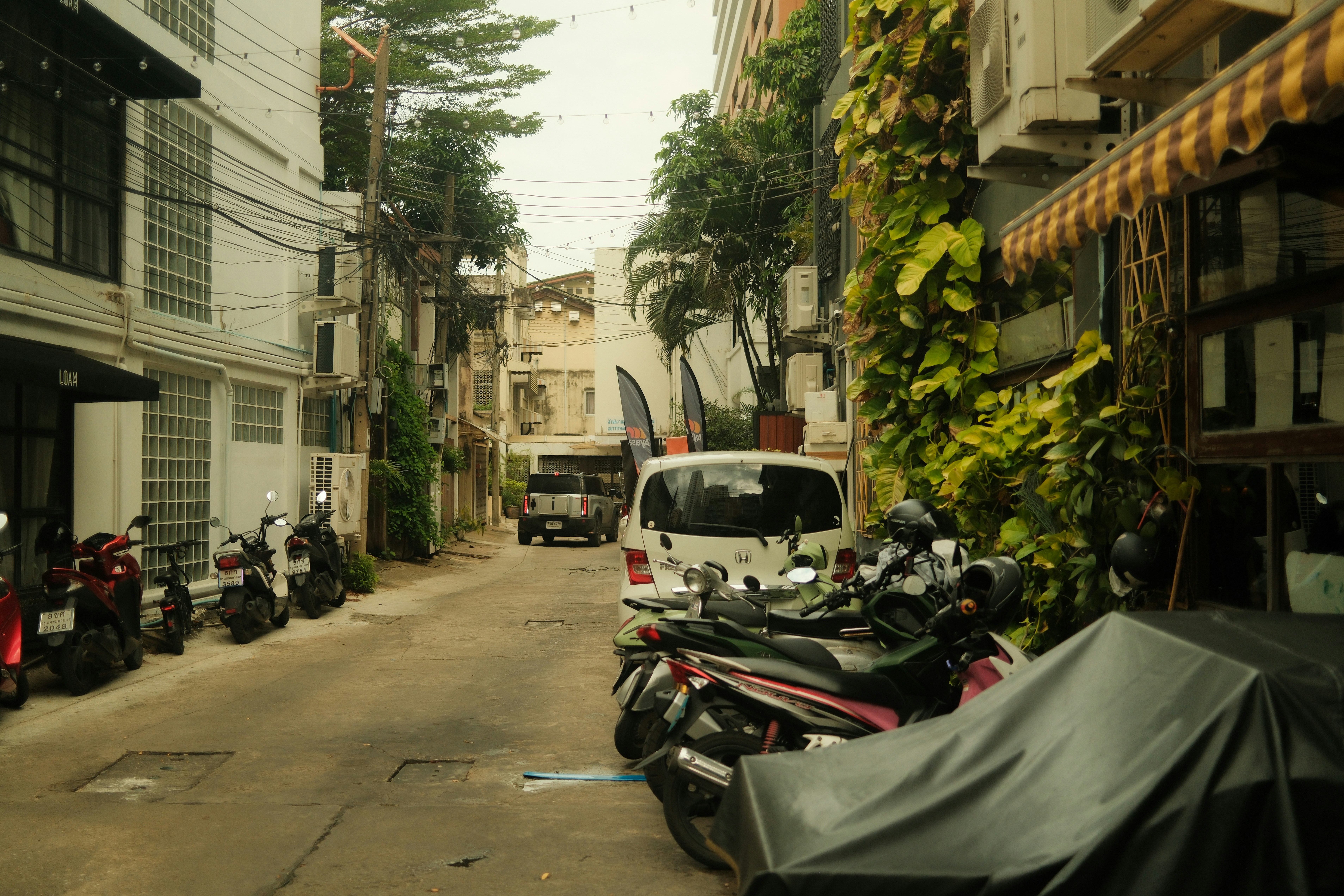 A narrow street lined with parked motorcycles and a white van drives toward the distance, framed by buildings and ivy-covered walls on the right. A calm, everyday urban scene with muted colors.