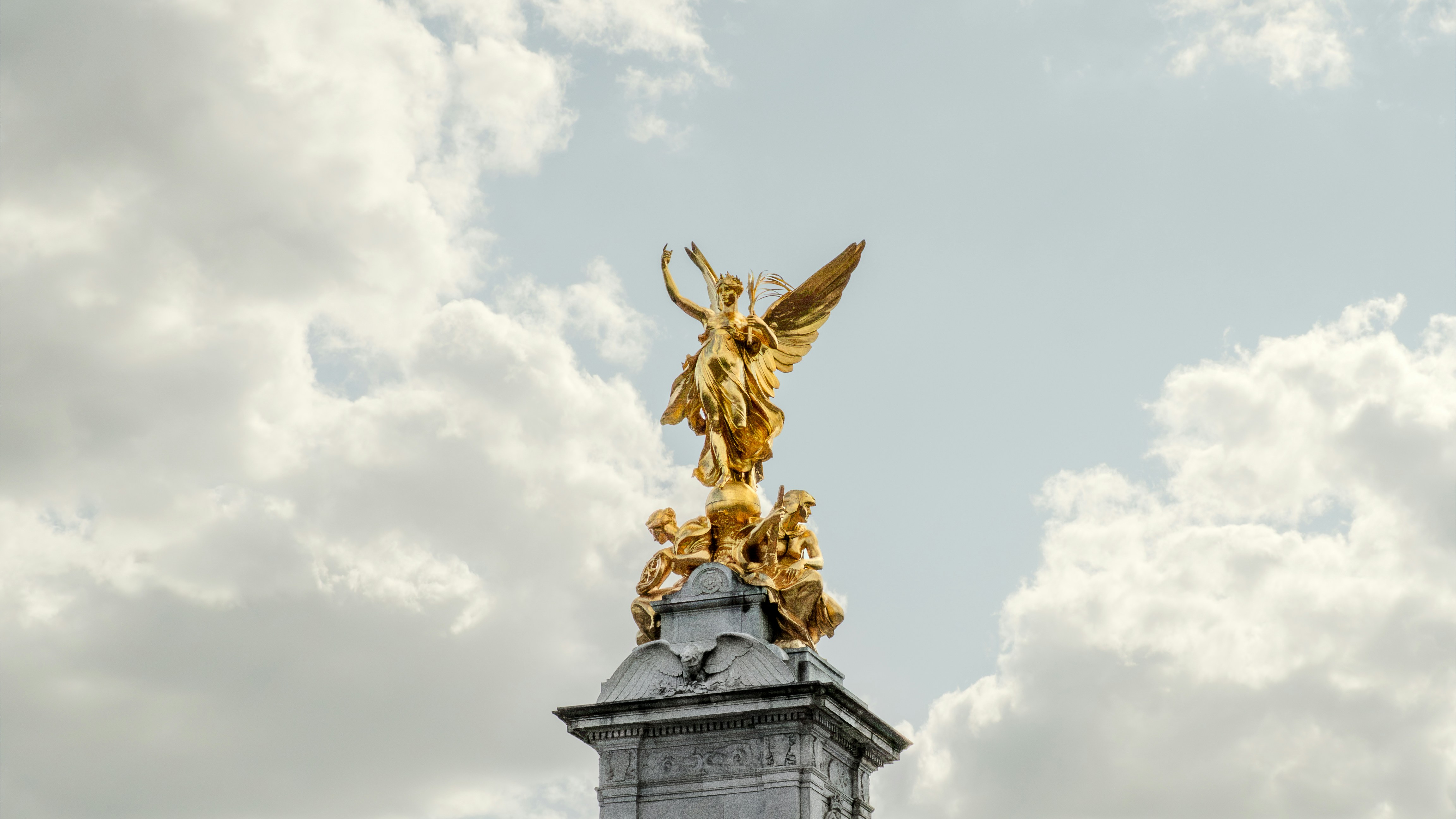 A gilded winged statue atop a dark pedestal against a bright sky; the statue stands as the focal point with surrounding clouds providing contrast.