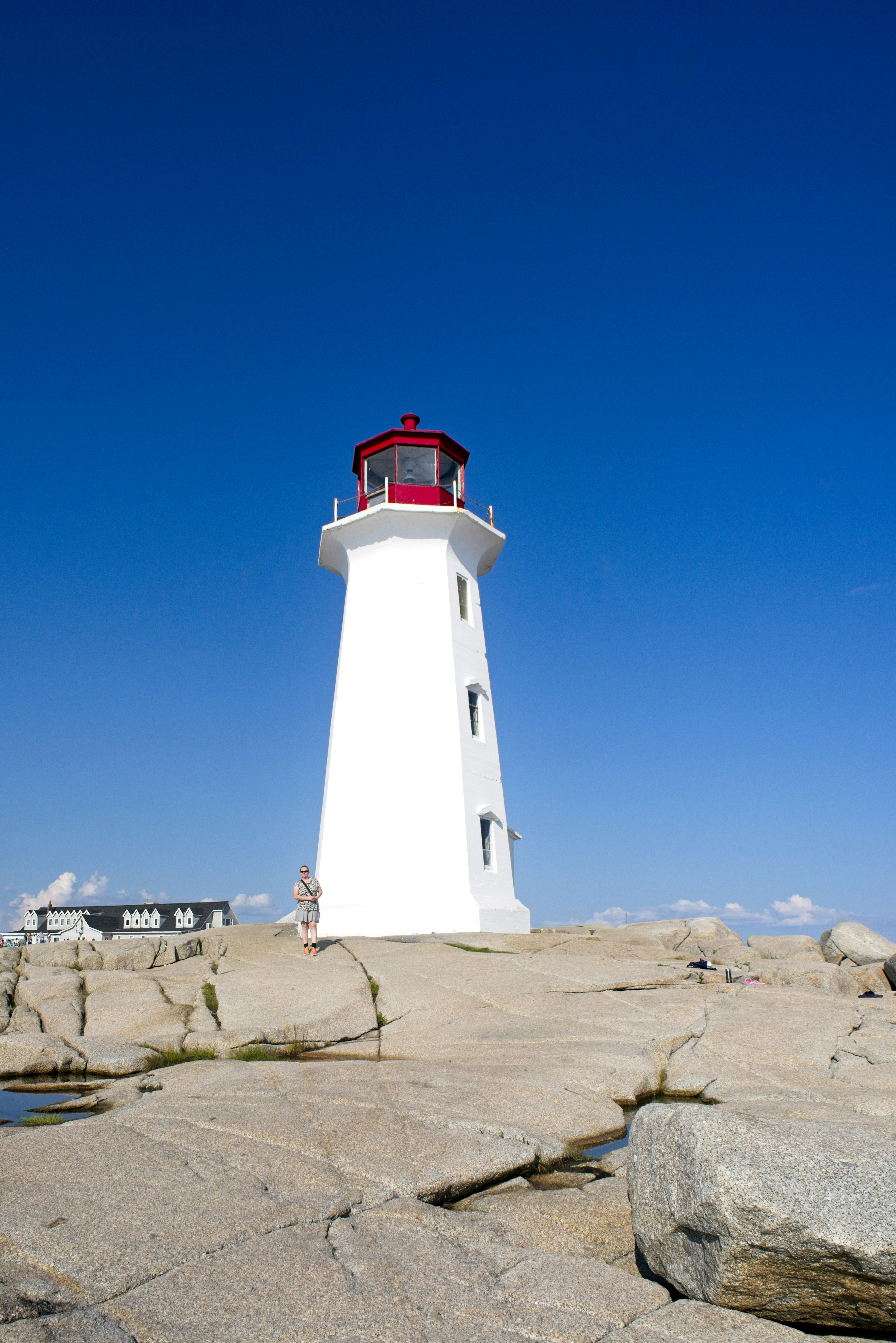 Standing proudly atop smooth, weathered granite, the famous Peggy's Cove Lighthouse gleams white against a deep blue sky on a sunny July afternoon. Its bright red lantern room crowns the octagonal tower, one of the most photographed landmarks in Atlantic Canada. A lone visitor pauses at its base, giving scale to the striking structure, while tidal pools glisten among the cracked glacial boulders in the foreground. In the distance, a white building with a dark roof sits along the rugged Nova Scotia coastline, completing this classic maritime scene.