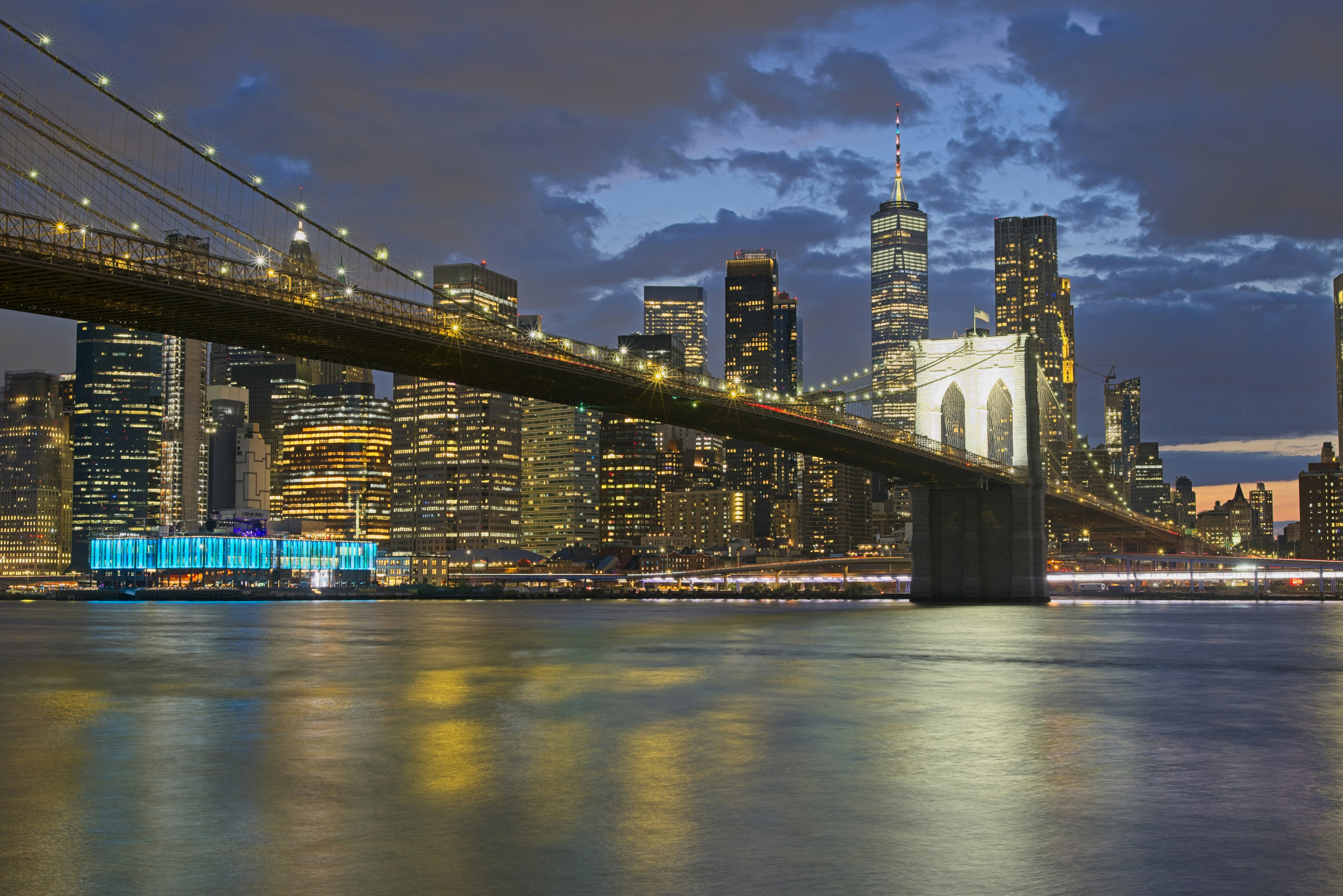 As dusk settles over New York on a hot July evening, the Brooklyn Bridge stretches gracefully across the East River toward a glittering Lower Manhattan skyline. Its illuminated Gothic stone tower and sparkling cable lights frame One World Trade Center, which rises proudly above the Financial District's glowing towers. Along the waterfront, Pier 17 shines in vivid turquoise, while a hint of orange sunset lingers on the horizon. A long exposure smooths the river into a silky mirror of golden and blue reflections, capturing the city's magical blue-hour atmosphere.