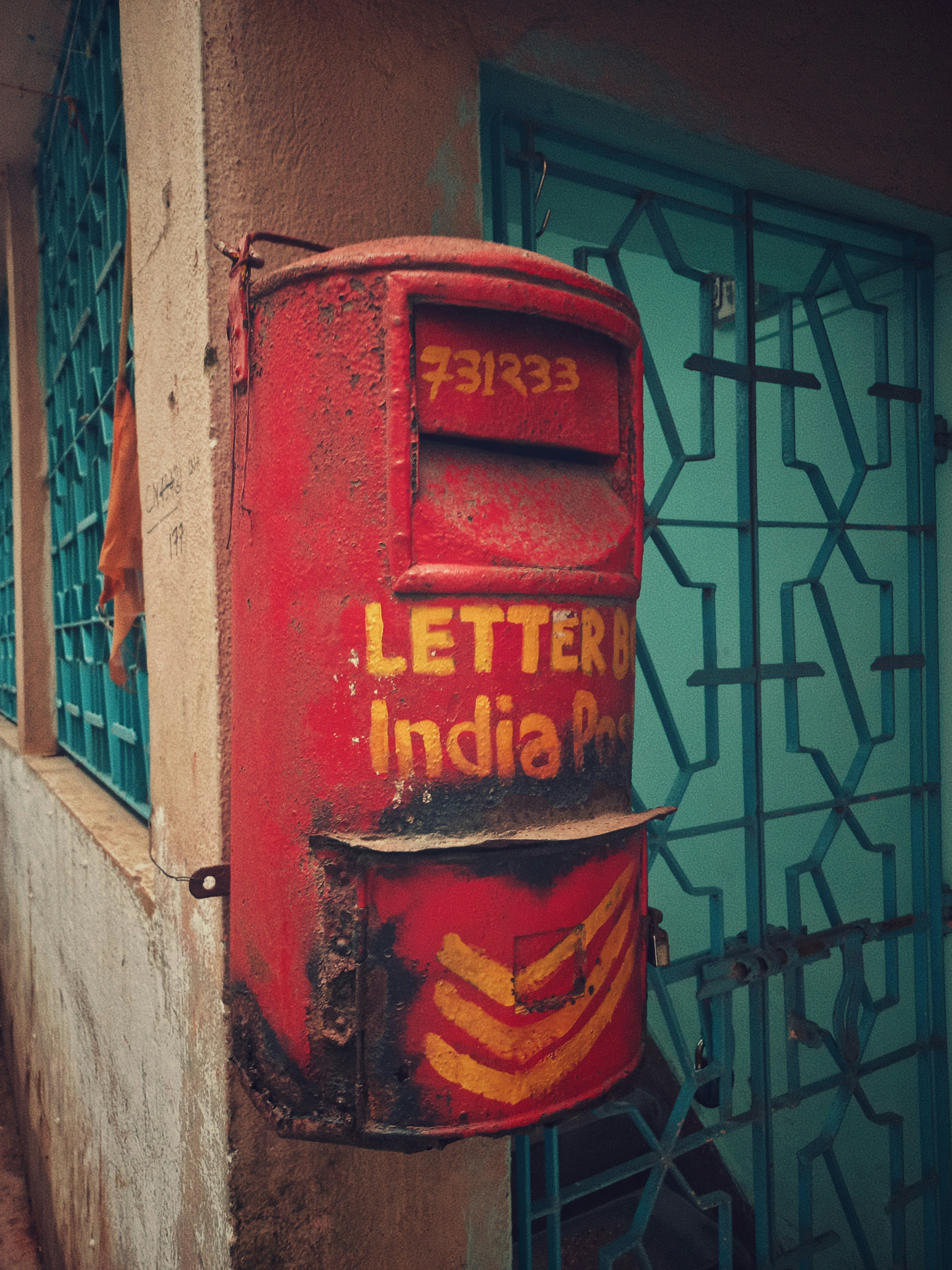 Weathered red postbox with faded yellow letters stands along a textured wall beside a teal metal gate.