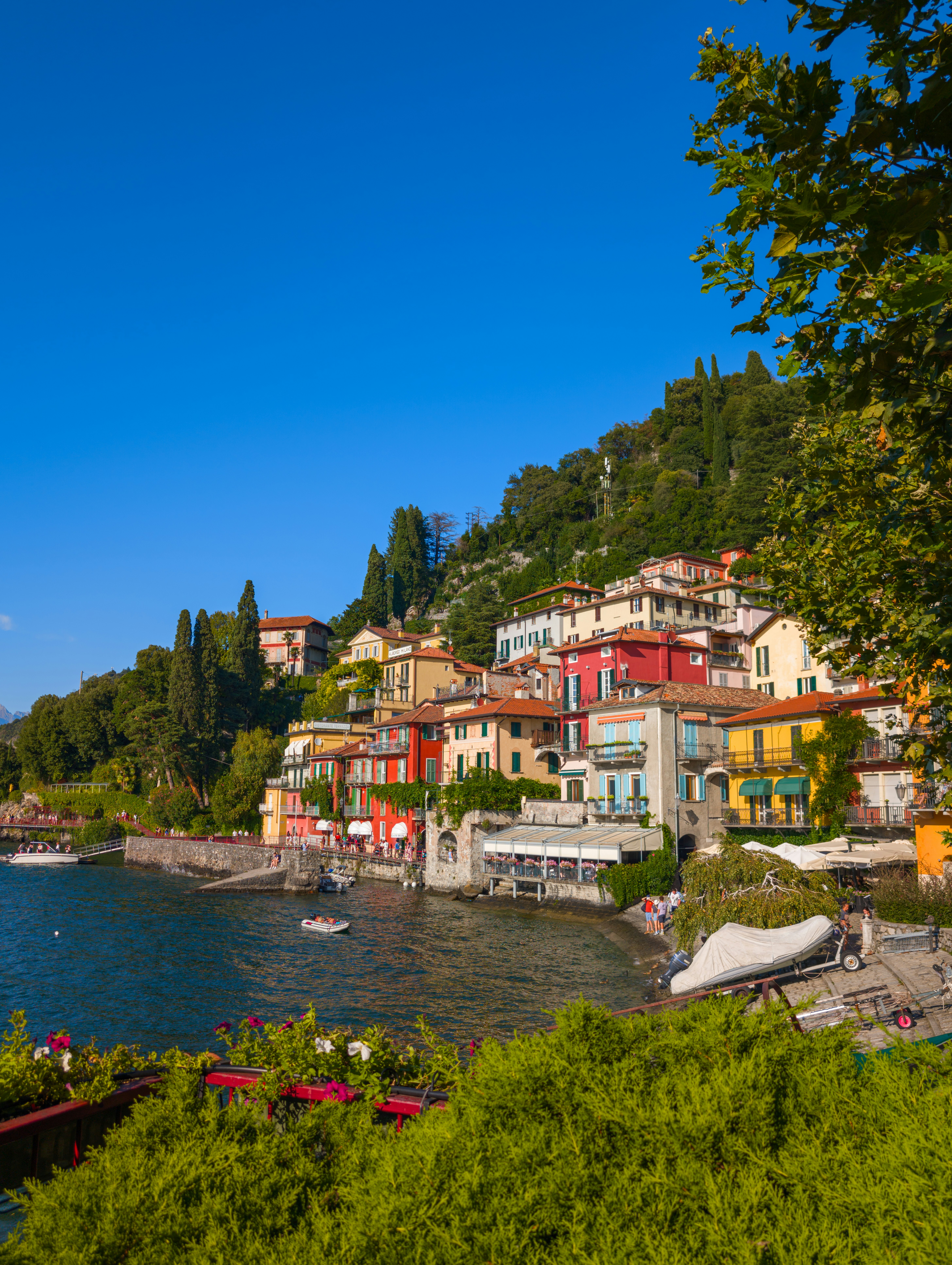 Colorful hillside village along a tranquil lake with boats and greenery. A sunny coastal town scene showcasing stacked houses and a stone quay, conveying a vibrant Mediterranean atmosphere.
