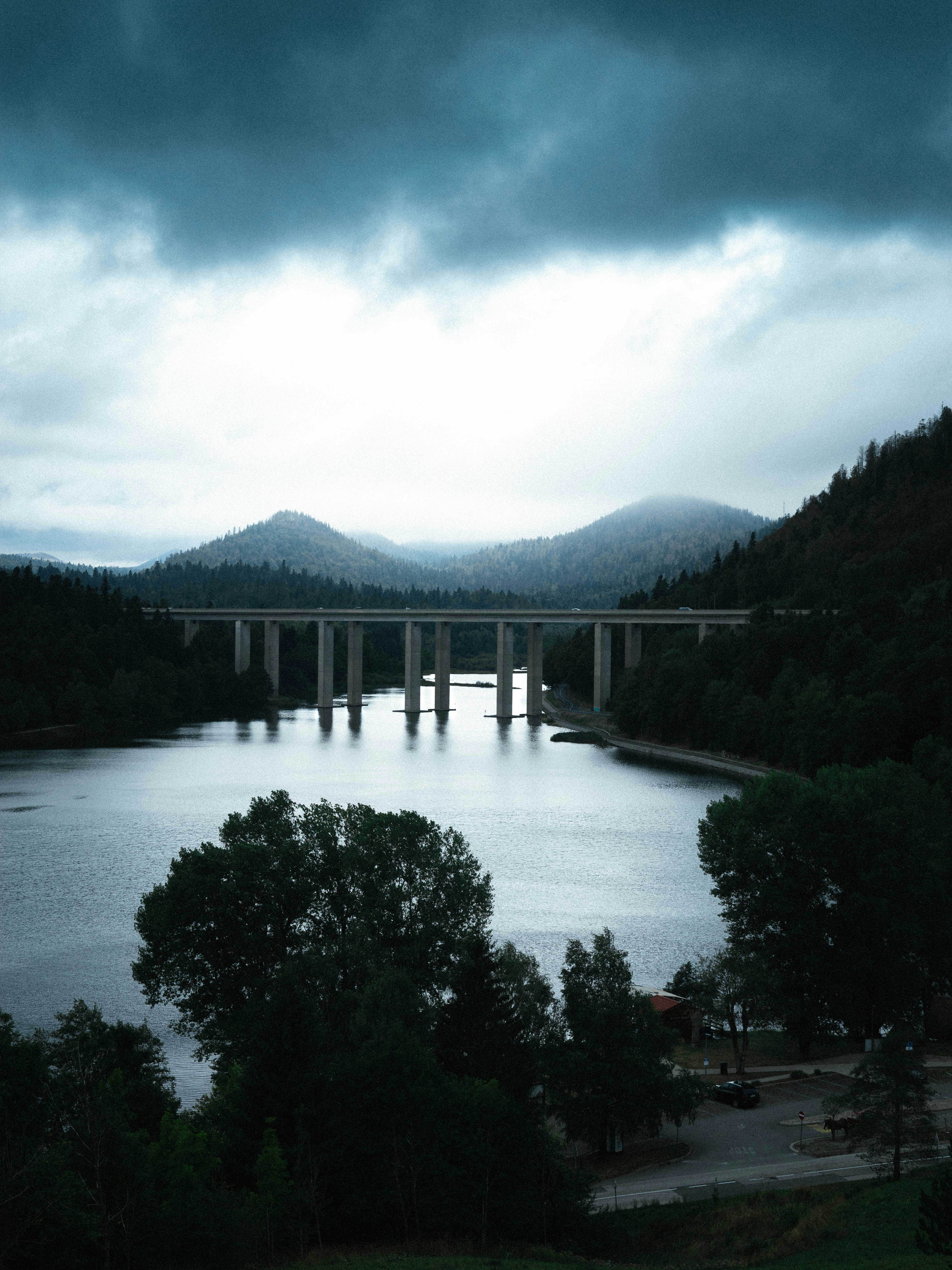 Reservoir with a distant bridge spanning calm water, framed by forested hills under a moody sky. The scene emphasizes the elevated viewpoint and serene urban-rural contrast.
