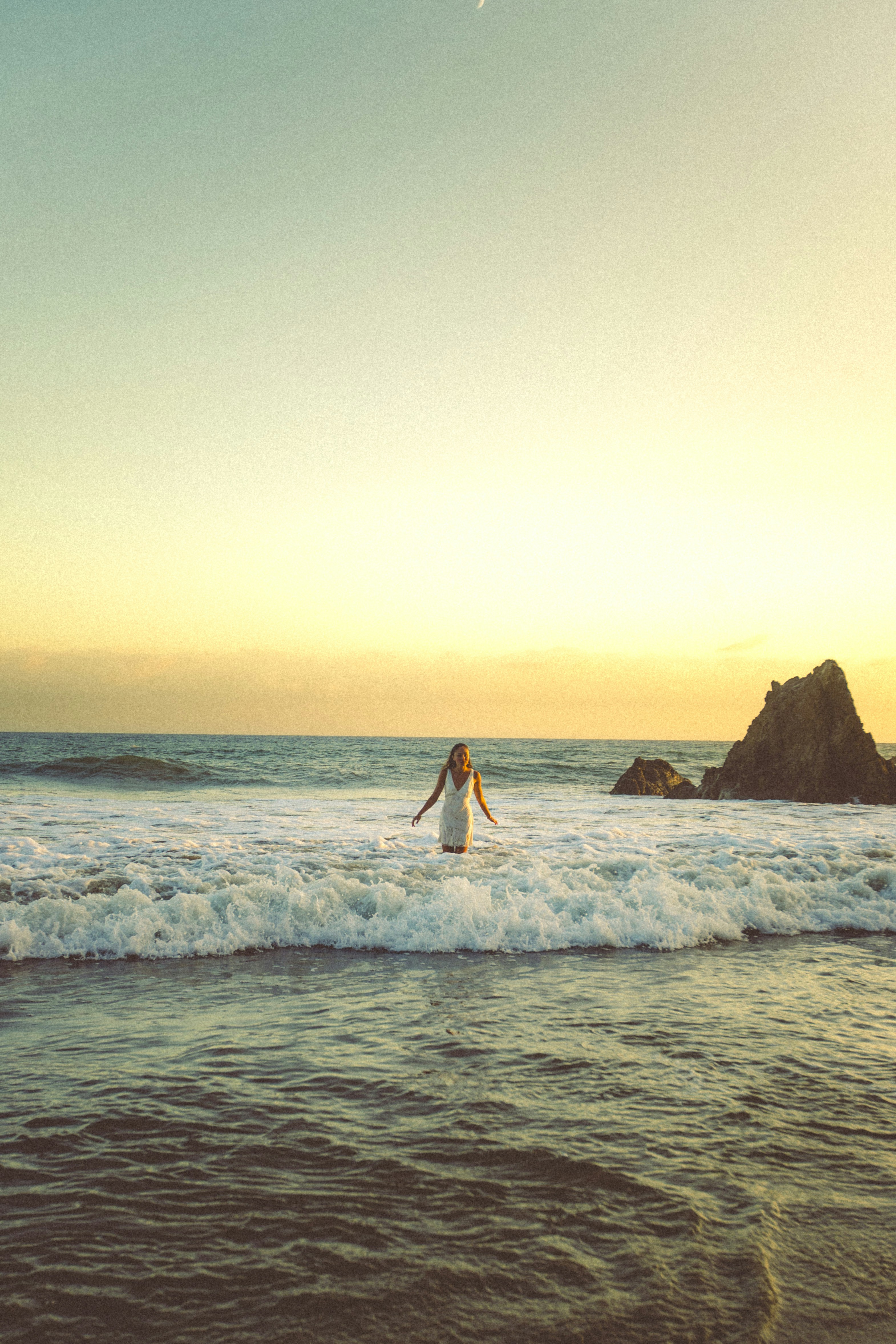 A lone figure stands waist-deep in gentle surf as sunlit waves roll in, with a rocky outcrop to the right and a warm, gradient sky above.