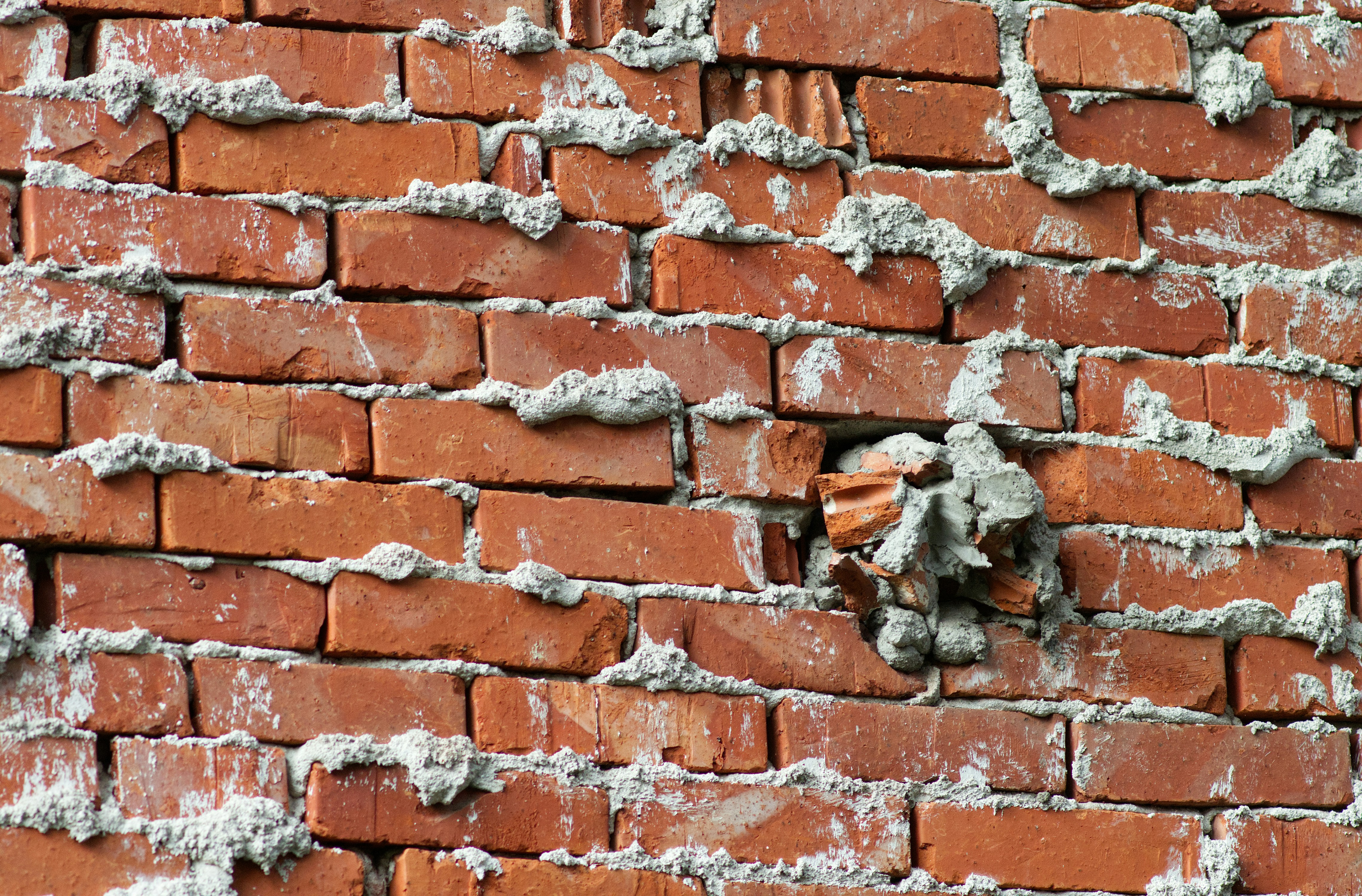 Red brick wall with irregularly poured concrete mortar and a small, weathered cavity near center-right. The texture emphasizes rough surface and urban decay; no people or animals present.