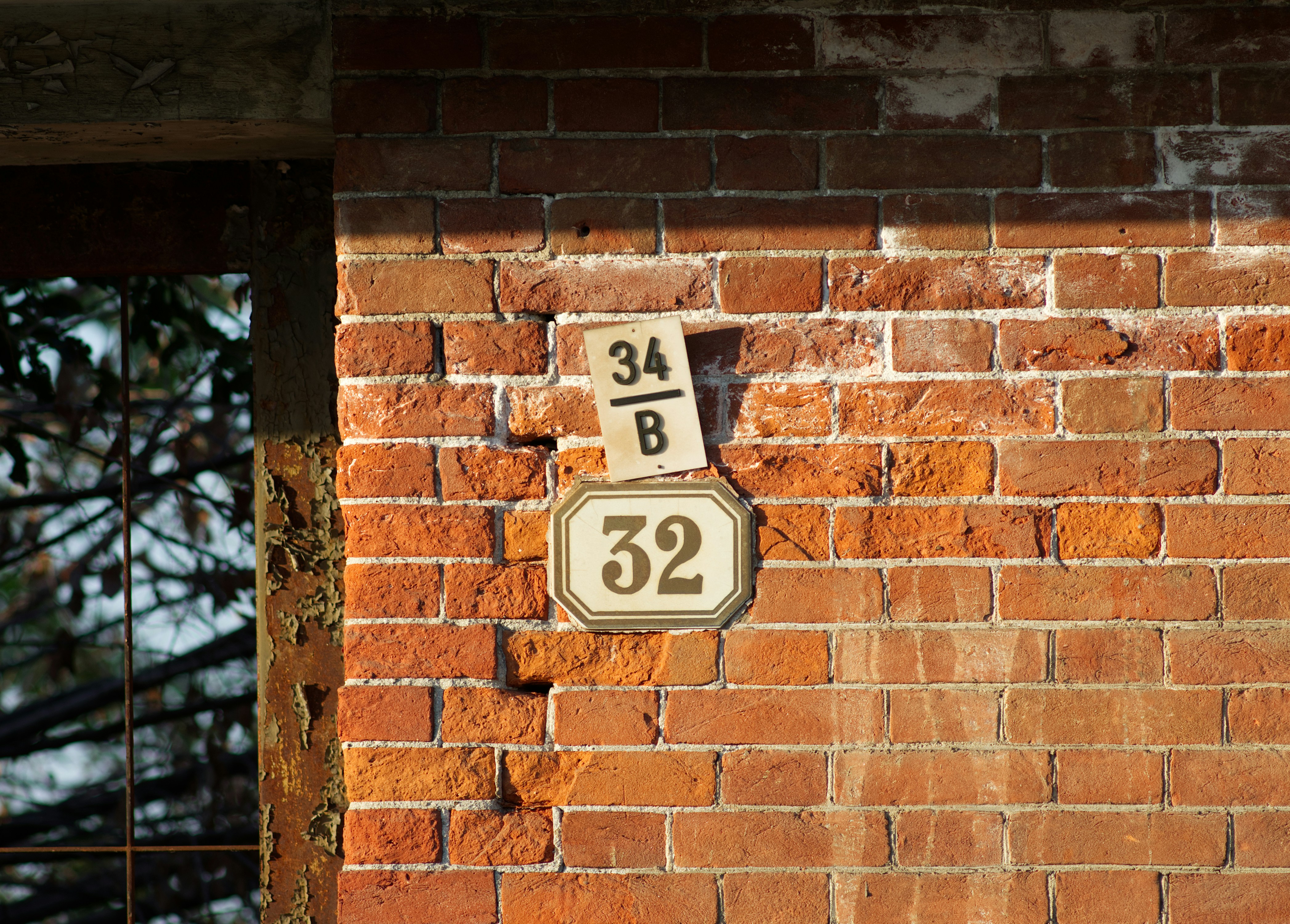 A weathered brick wall with a vintage address plaque (32) and a smaller sign (34 B) above it, captured with warm afternoon light showing texture and shadows on the aged surface.