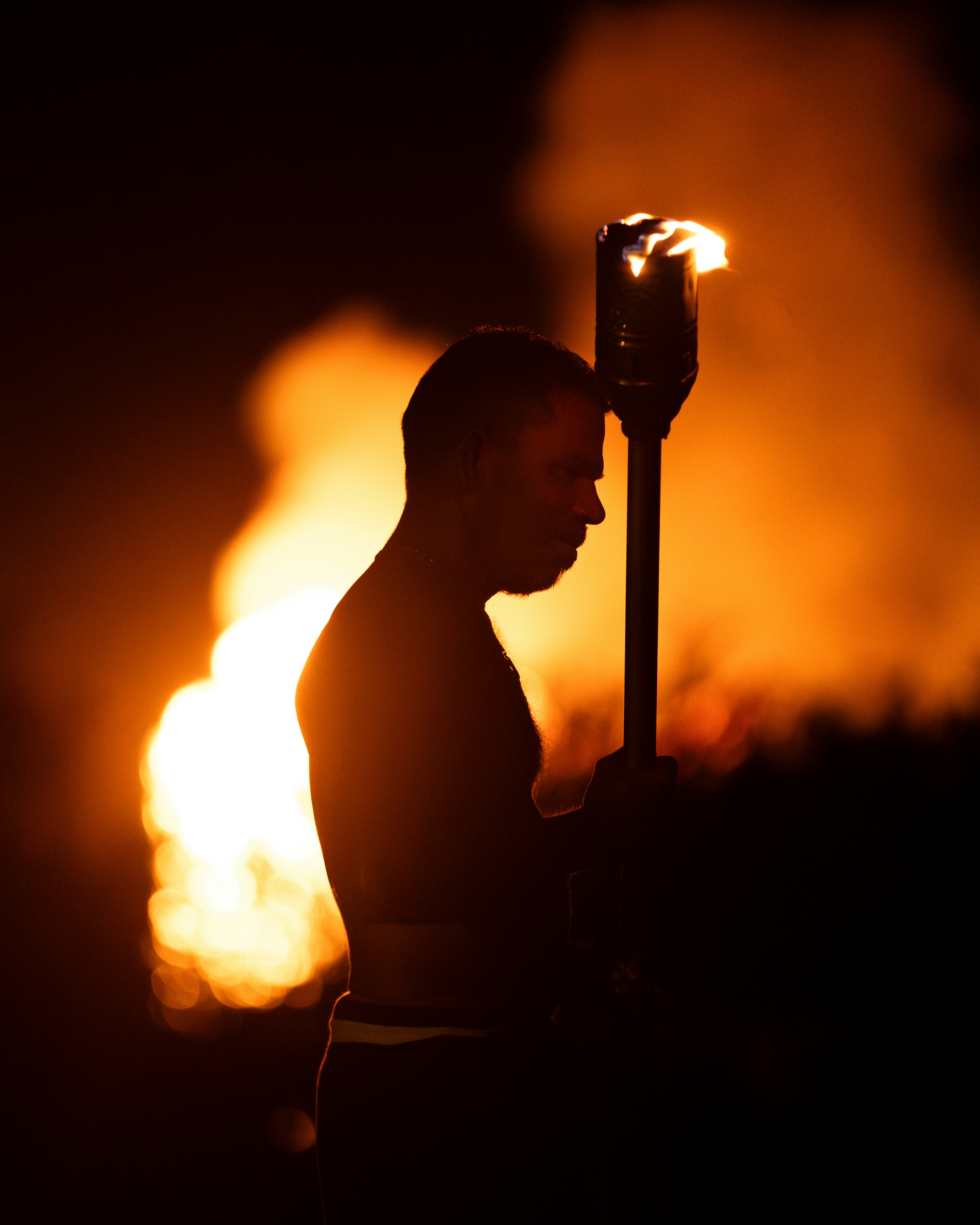 Silhouette of a person holding a torch against a blazing backdrop, with dramatic orange flames creating a backlit scene. This appears to be a live-action capture, likely documenting a fire-lit performance or ritual.