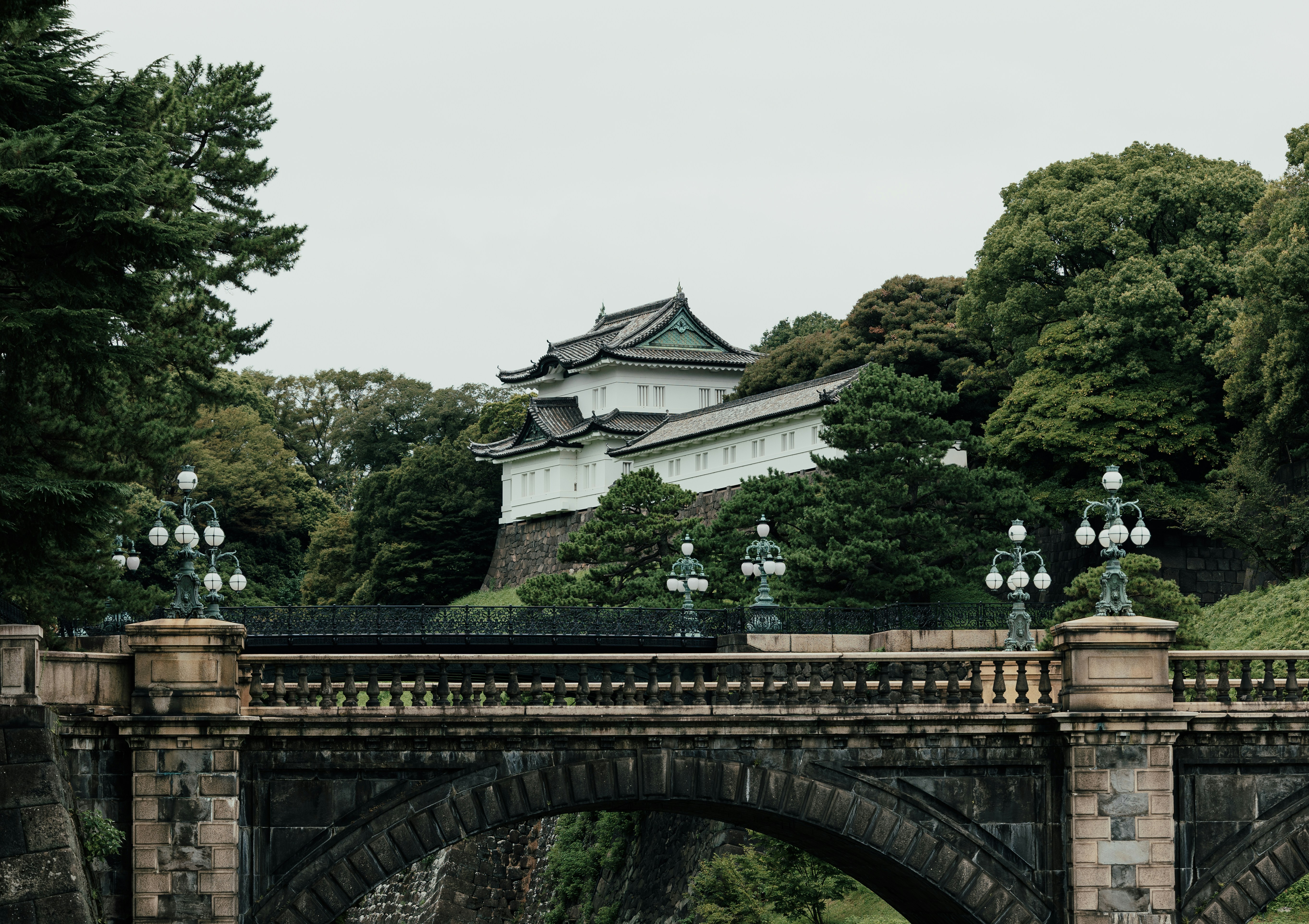Stone bridge and traditional Japanese castle sit above a calm river with lush trees in the background, presenting a tranquil architectural scene.