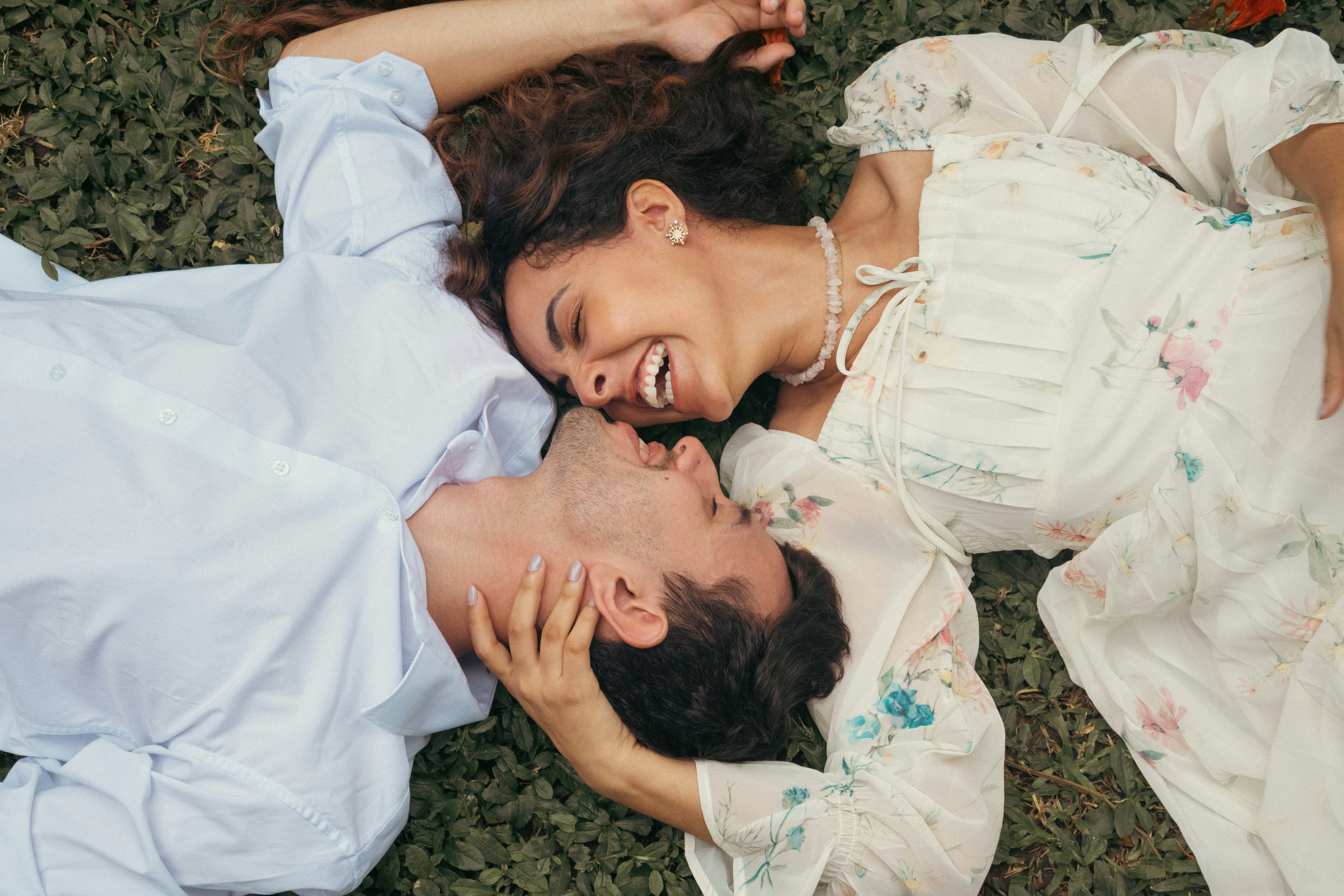 Two lovers lie on the ground, facing each other with smiles, in a light floral dress and a pale blue shirt; a tender moment captured outdoors among greenery.
