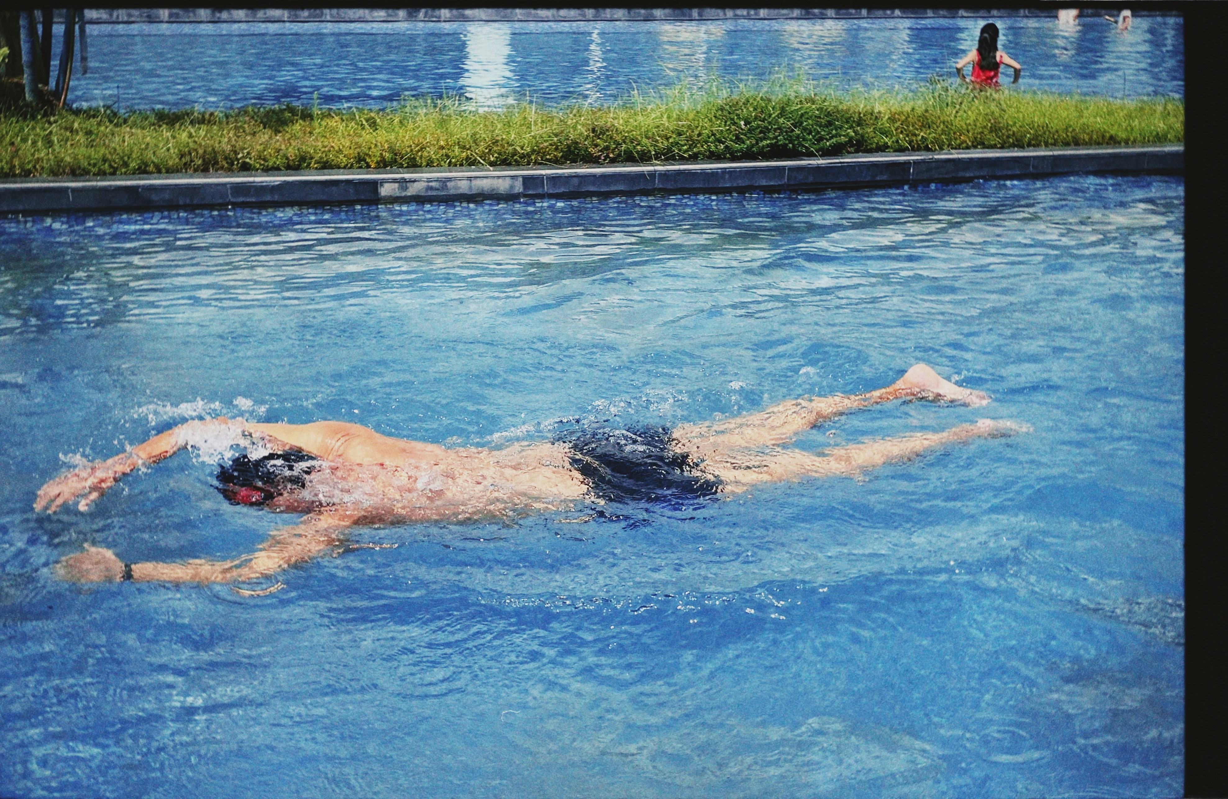 A swimmer cuts through clear blue water in a sunlit pool, with the far grassy edge and a person at the poolside visible in the background. This is a photograph capturing a swimmer in mid-stroke near the surface.