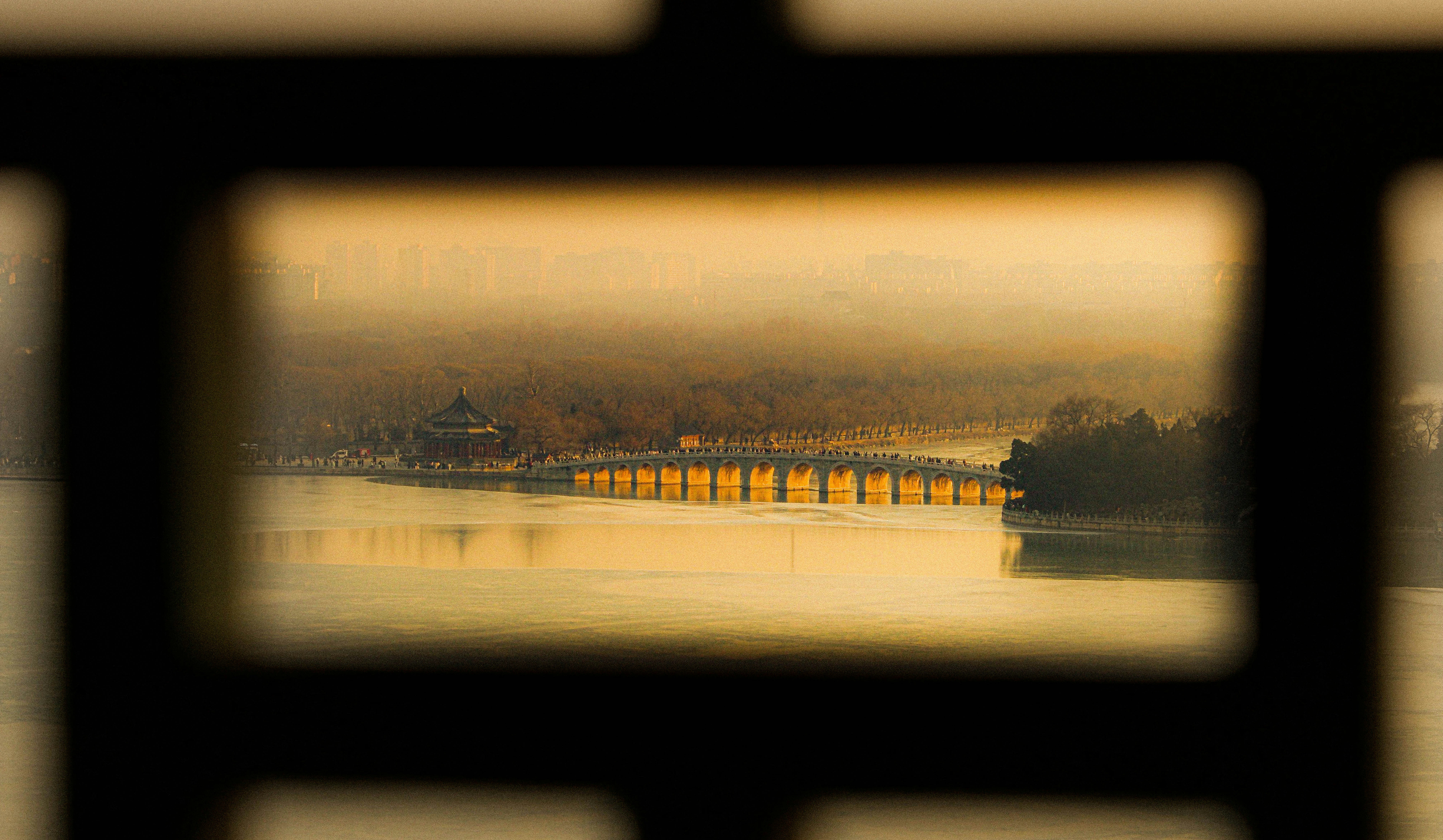 Sunset scene peeks through dark window frames to reveal a golden bridge over calm water.