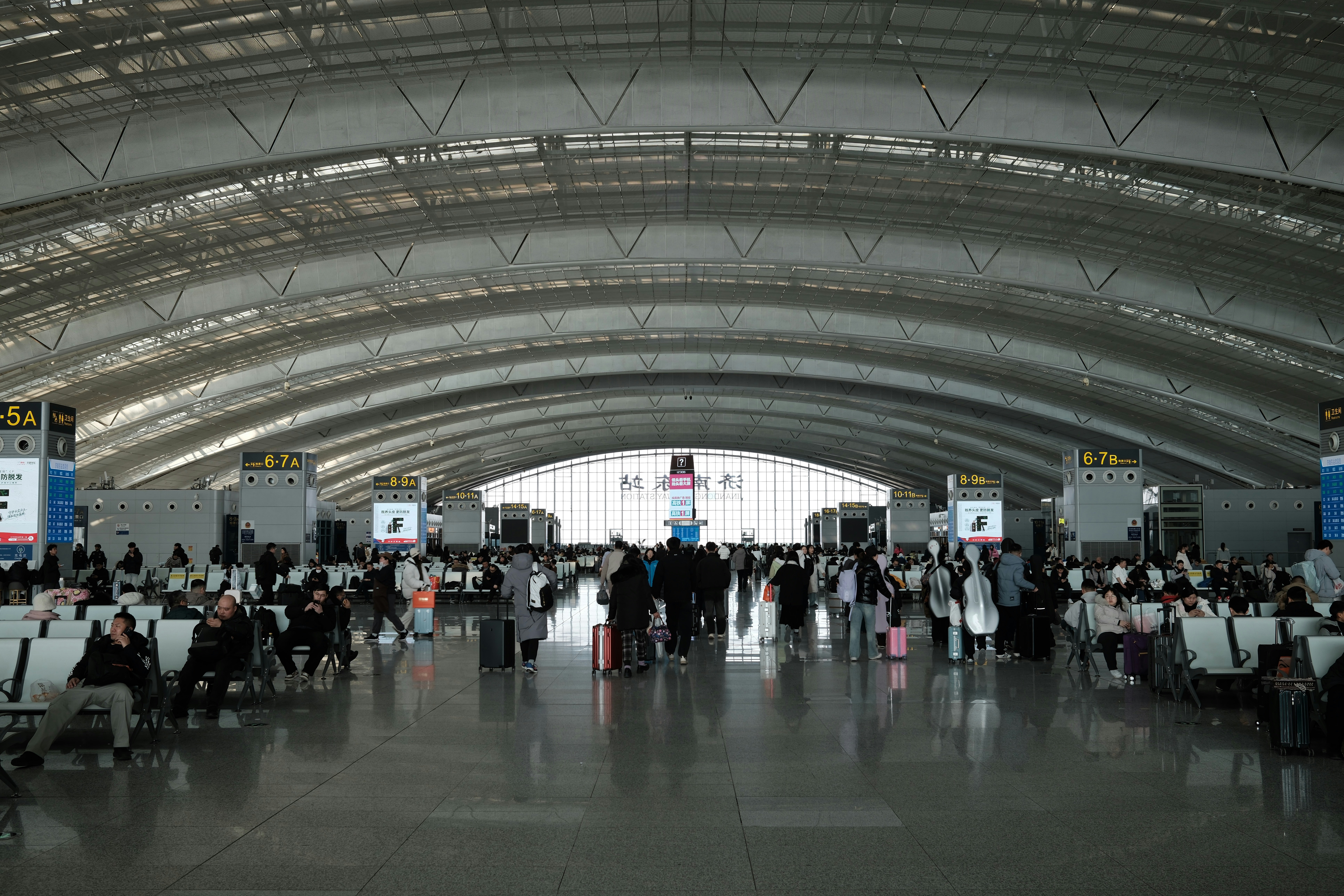 Busy airport terminal with travelers seating and queues under a curved glass roof; signage and distant departure boards mark a high-traffic concourse.