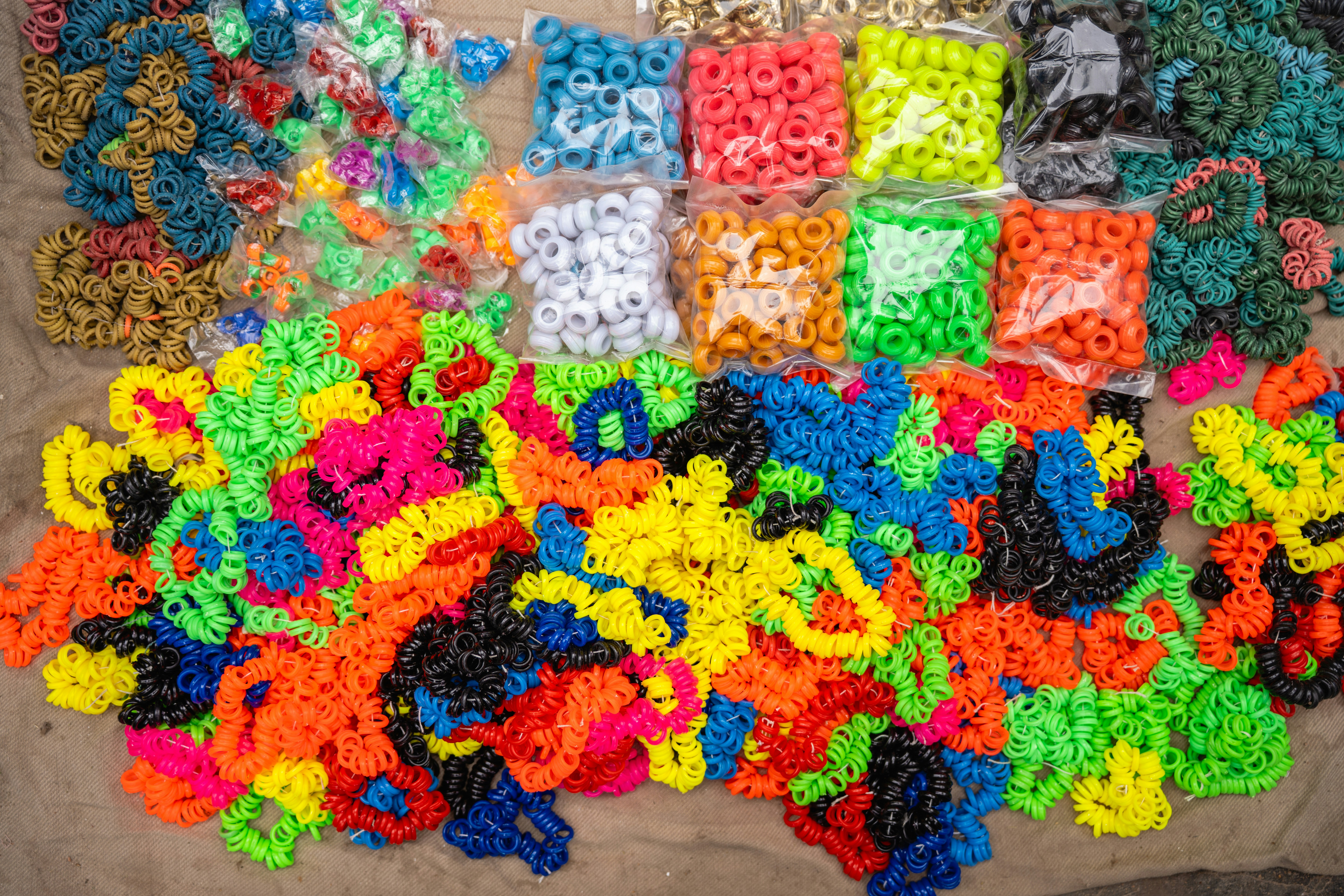 Vibrant bundles of curled plastic cords arranged in a market display, with additional bins of assorted colors at the top; a vivid craft stall scene.