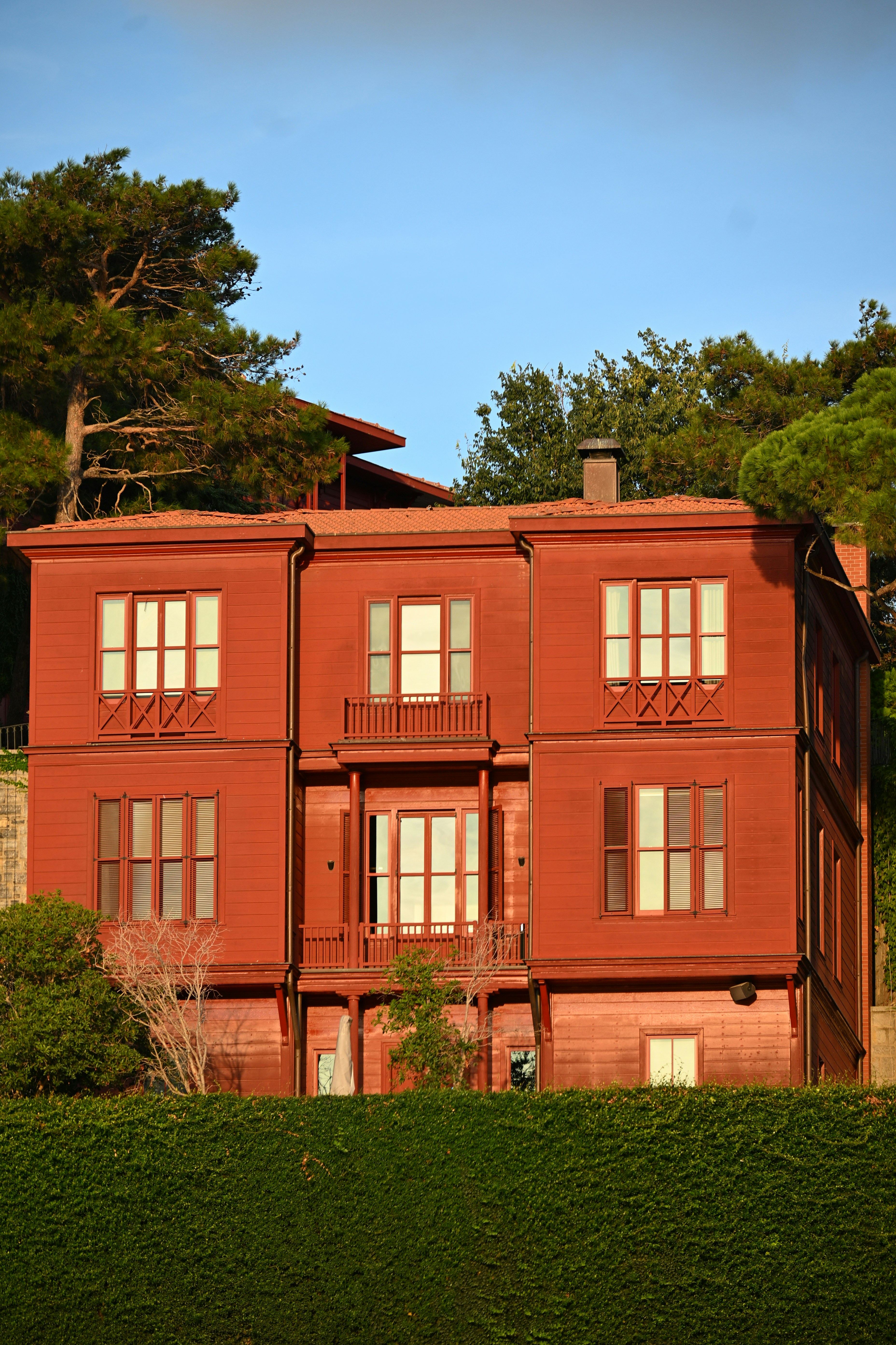 Sunlit red-brick villa with white-framed windows sits behind a trimmed green hedge under a clear blue sky.