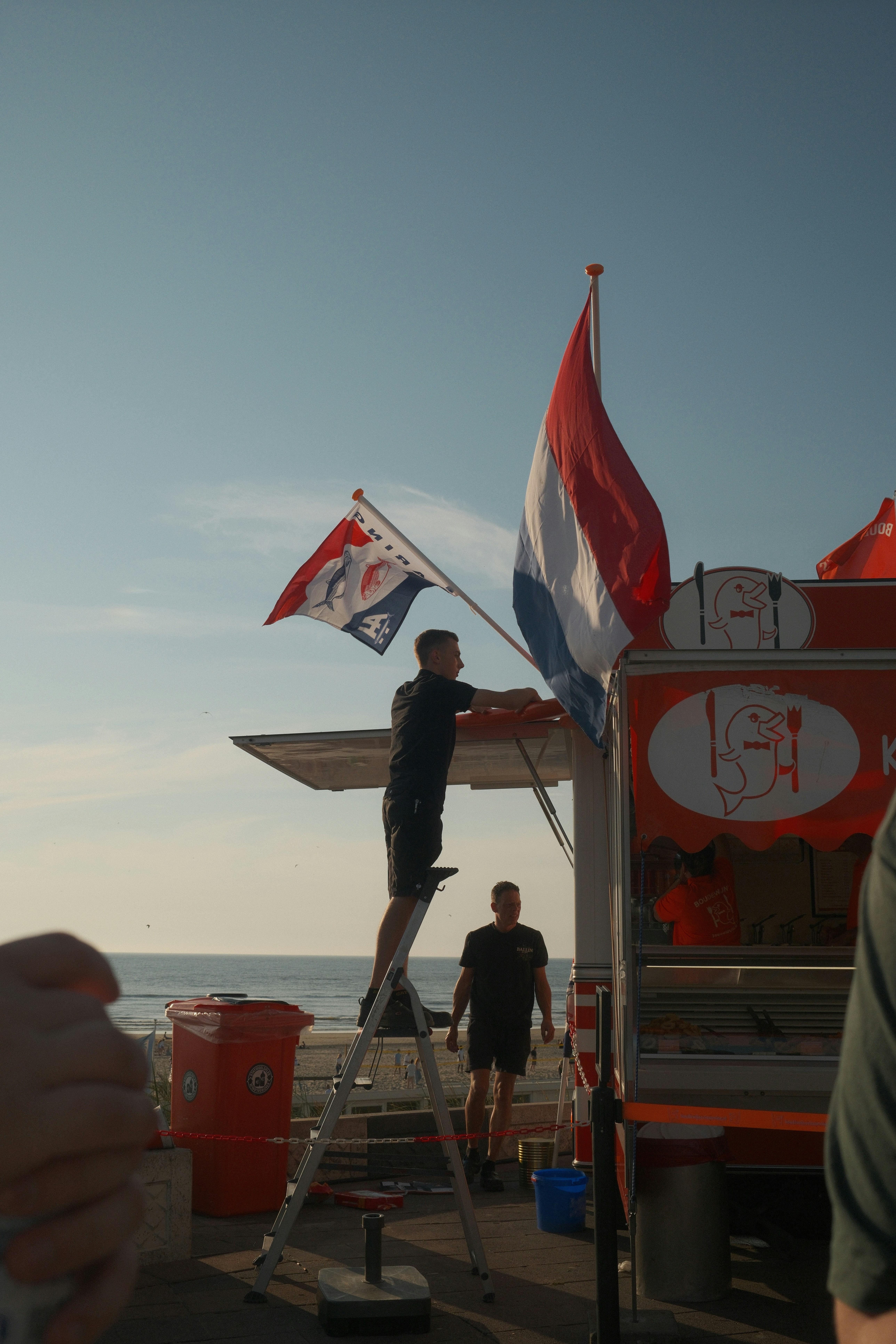 A person on a stepladder adjusting a Dutch flag on a food stall