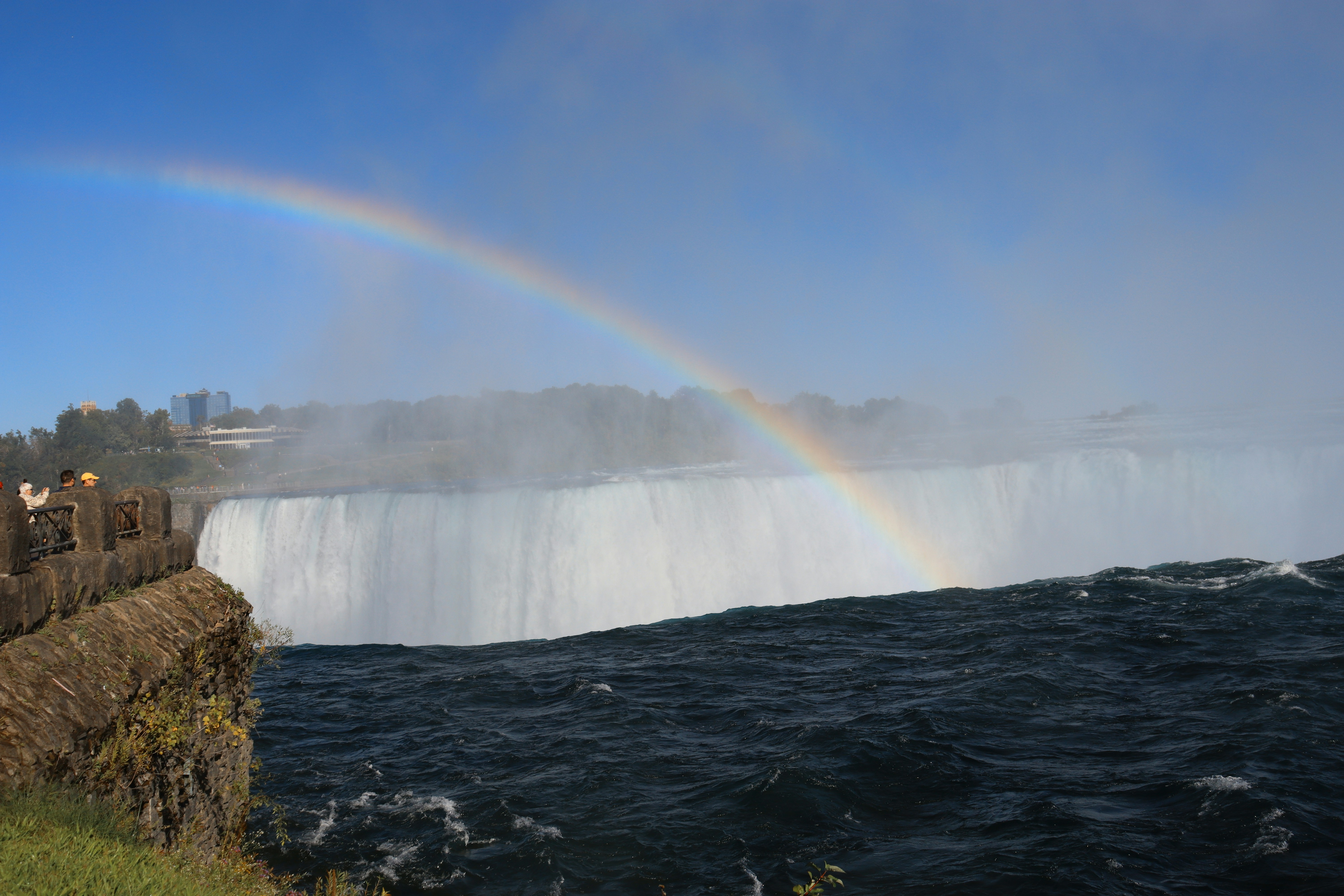 Rainbow arcing over the misty waterfall edge with a rocky ledge in the foreground and distant skyline; a landscape shot capturing the spray and color near a waterfall.