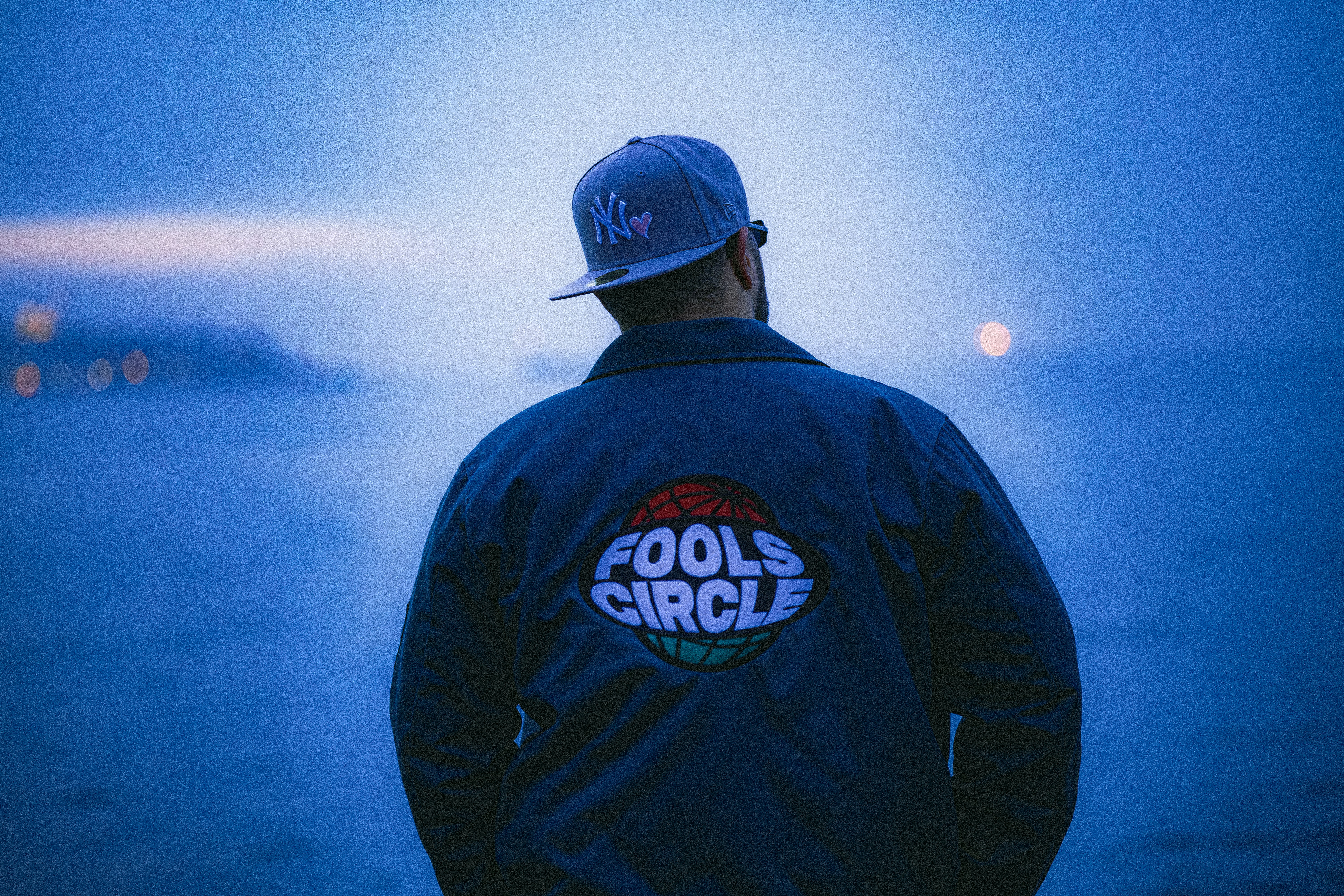 Back view of a person wearing a dark jacket with a circular 'FOOLS CIRCLE' logo, standing near a blue-tinted body of water at dusk.