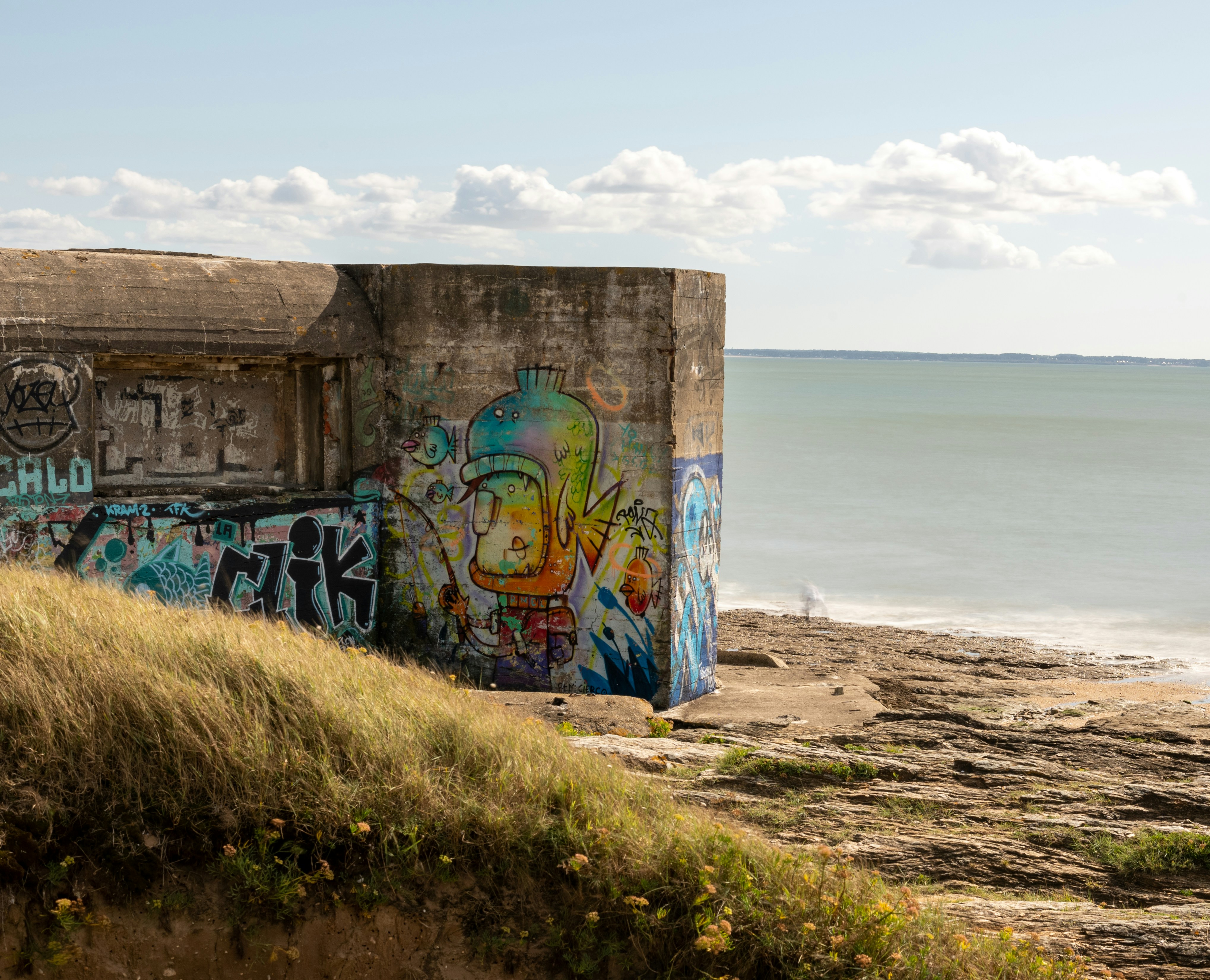 Graffiti-covered concrete structure by the sea at low tide, with grass foreground and a calm horizon.