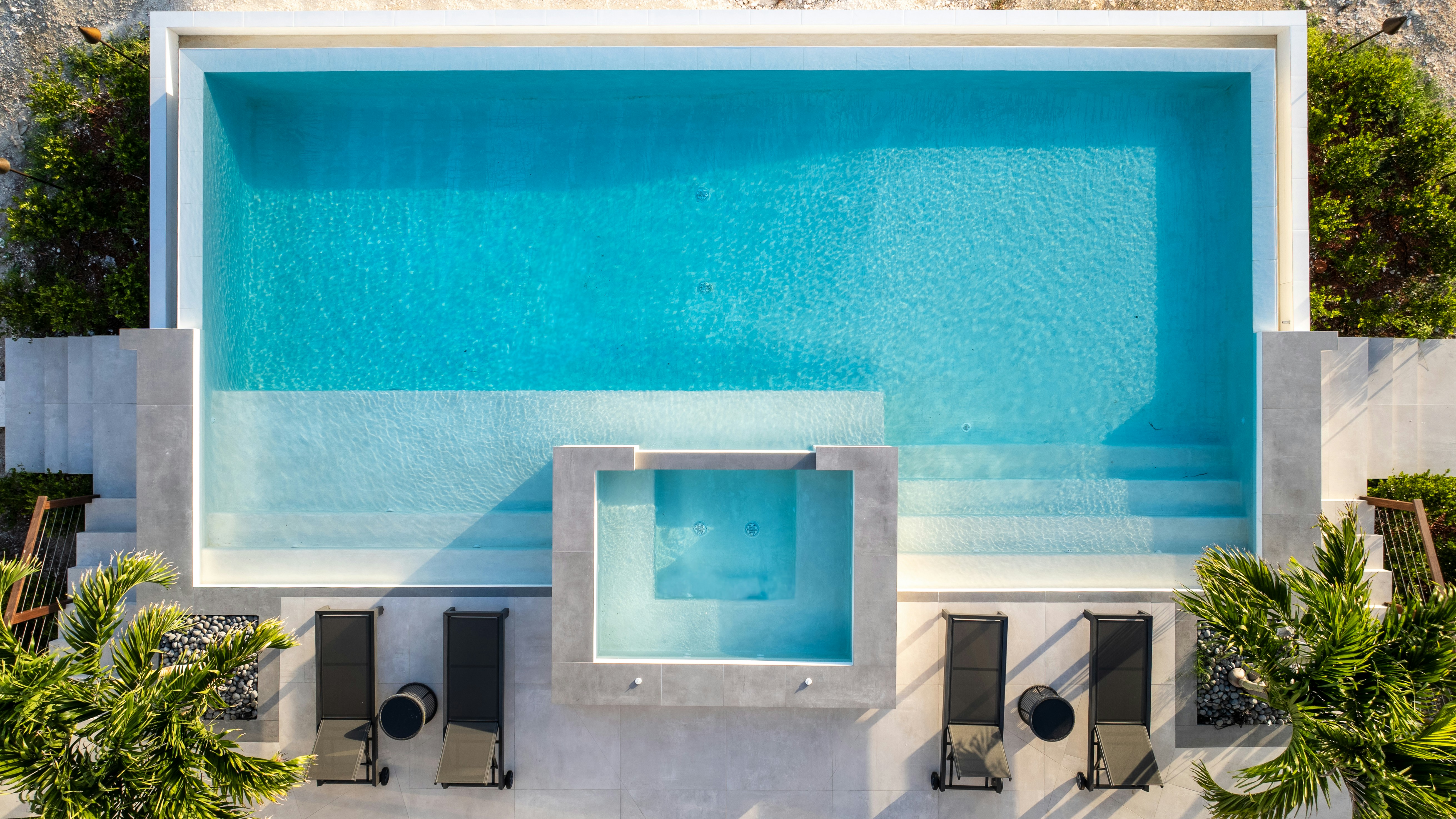 Aerial view of a pristine turquoise pool with an inset square spa, framed by a modern concrete deck and lounge chairs in a tropical setting.
