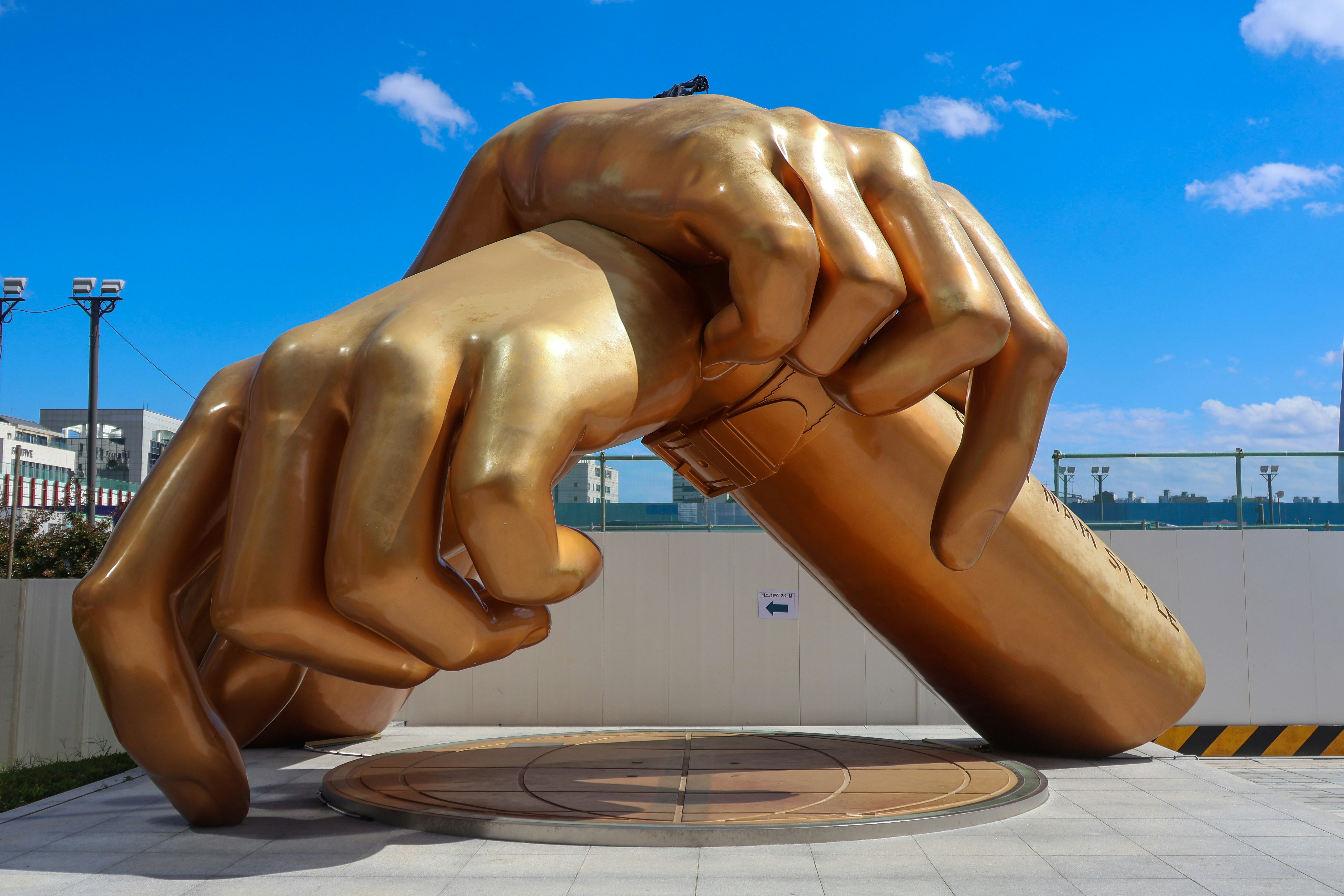 Golden sculpture of intertwined oversized hands outdoors with a clear blue sky; a ceremonial plaza setting suggests public art installation.