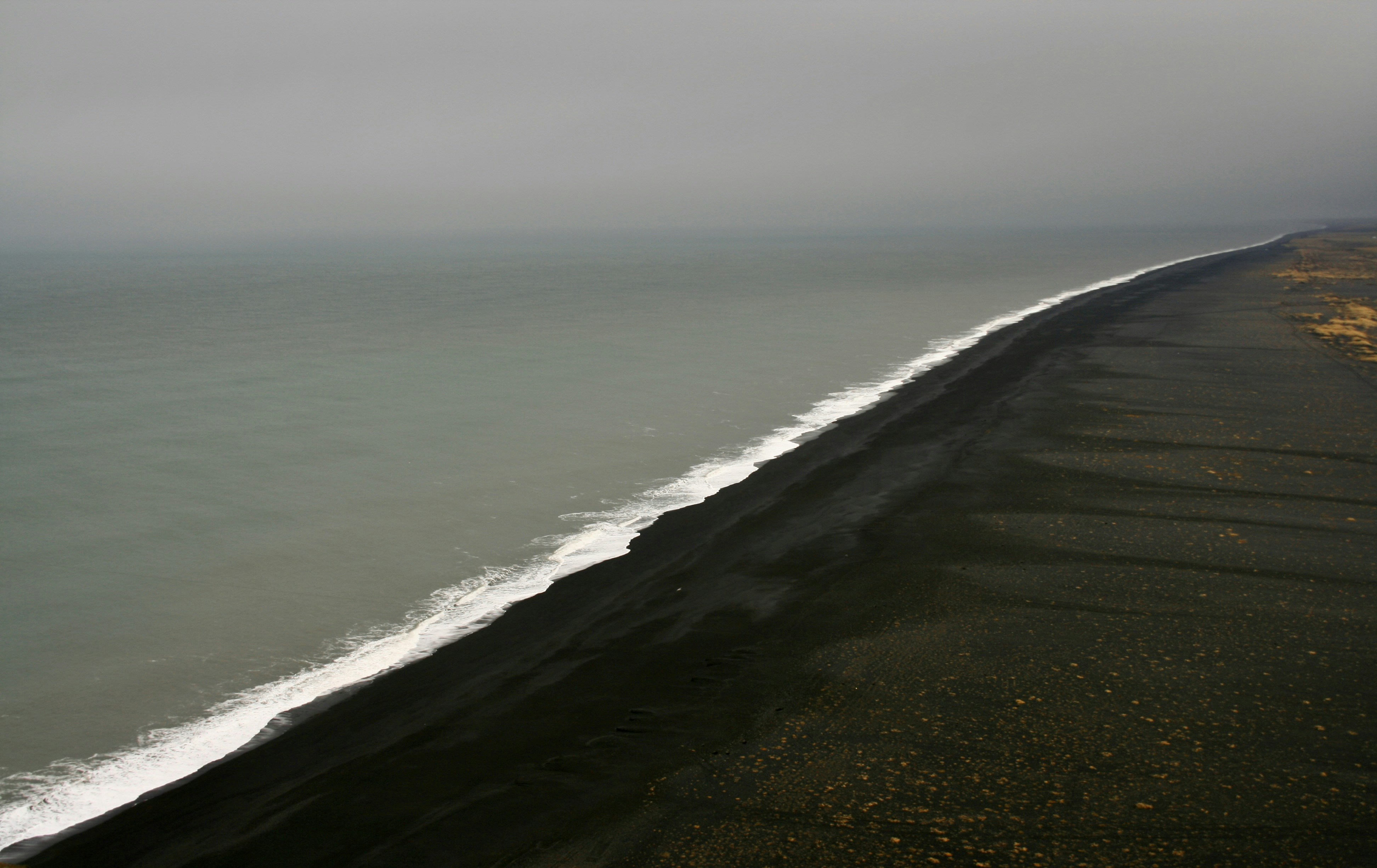 Dark shoreline curves toward the horizon with a pale, misty sea and a narrow strip of land on the right; the image emphasizes the contrast between the black beach and the pale ocean.