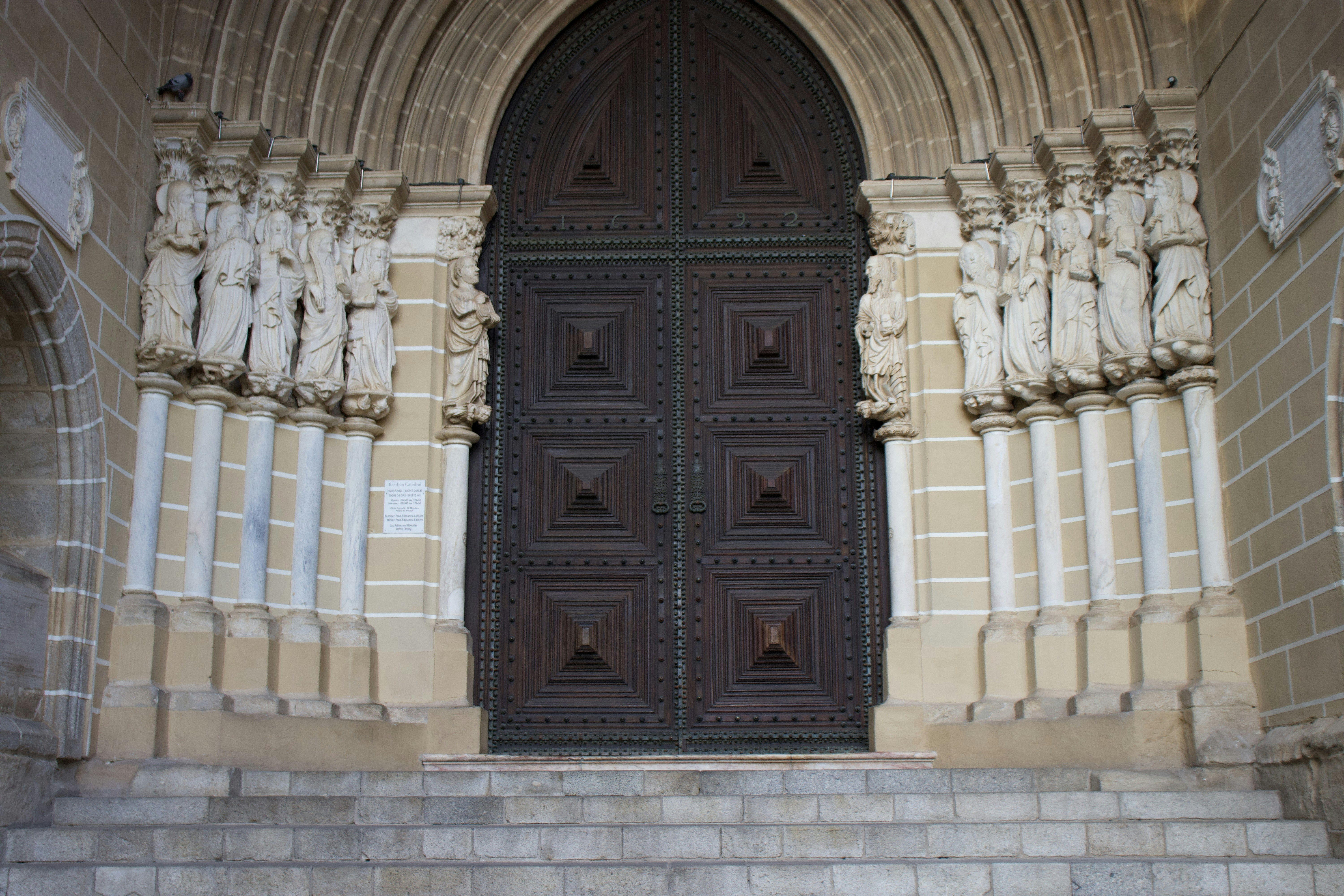 Intricately carved stone saints flank a heavy wooden door set in a vaulted arched entrance. The steps and symmetrical figures frame the central doorway, emphasizing architectural detail and stonework.