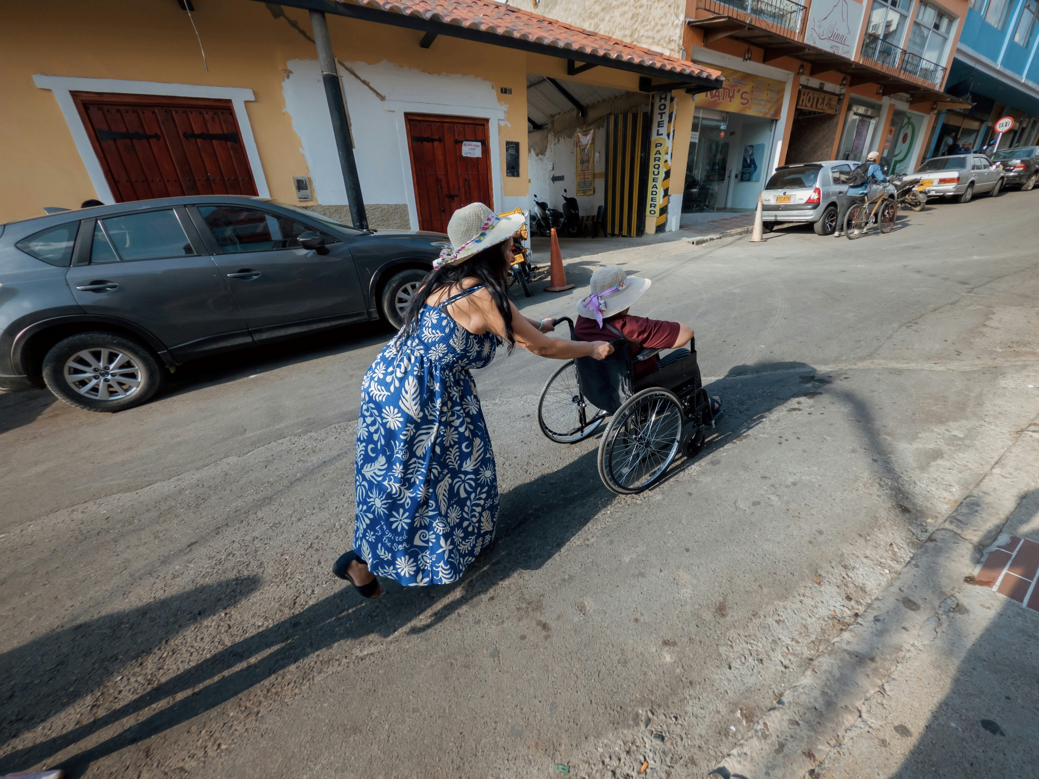 Sunlit Exchange: Woman in a Blue Dress Guides an Elderly Rider Along a Sun-Warmed Street