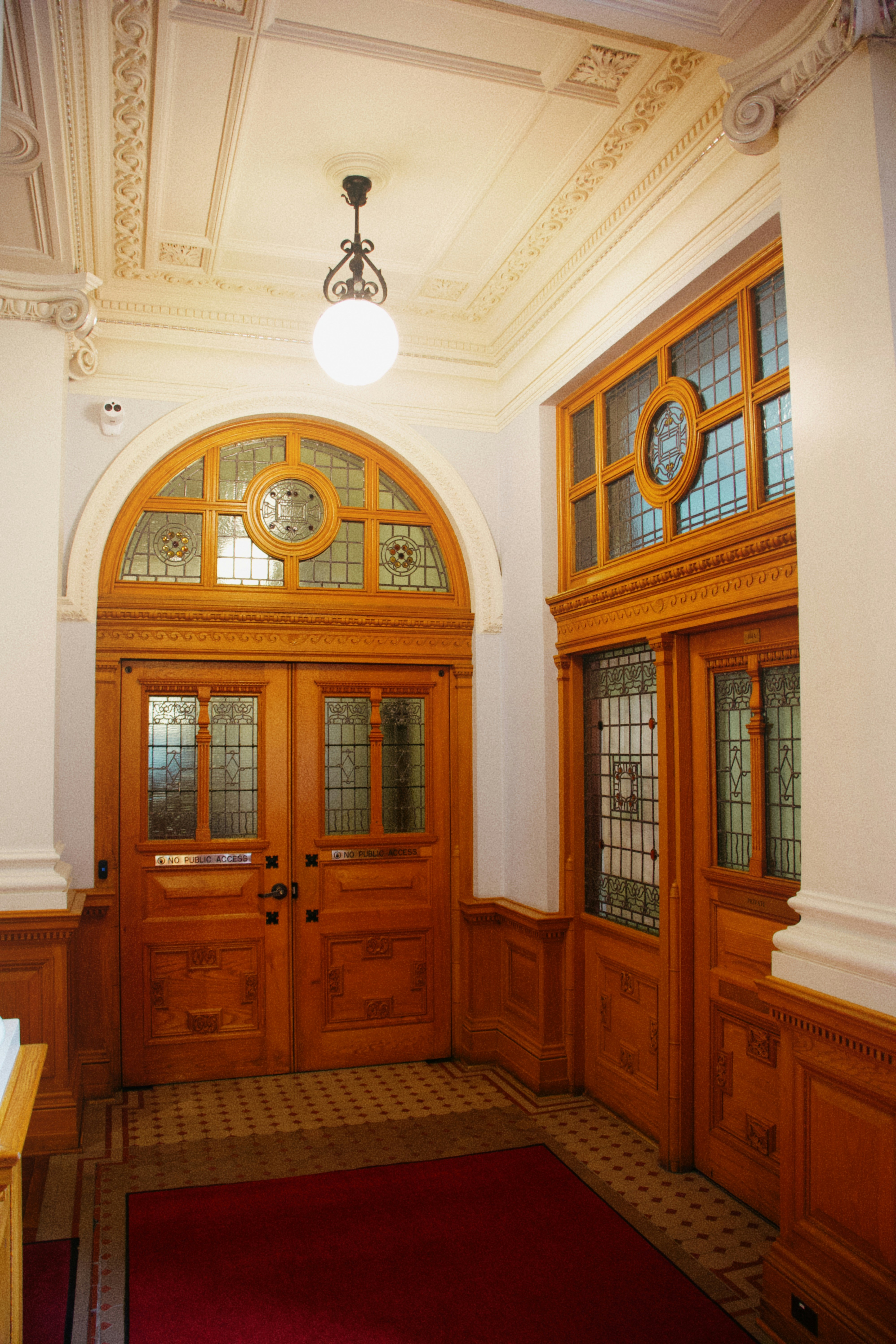 Ornate wooden doors with stained glass panels in a grand foyer; warm lighting and patterned tile floor frame the entryway.