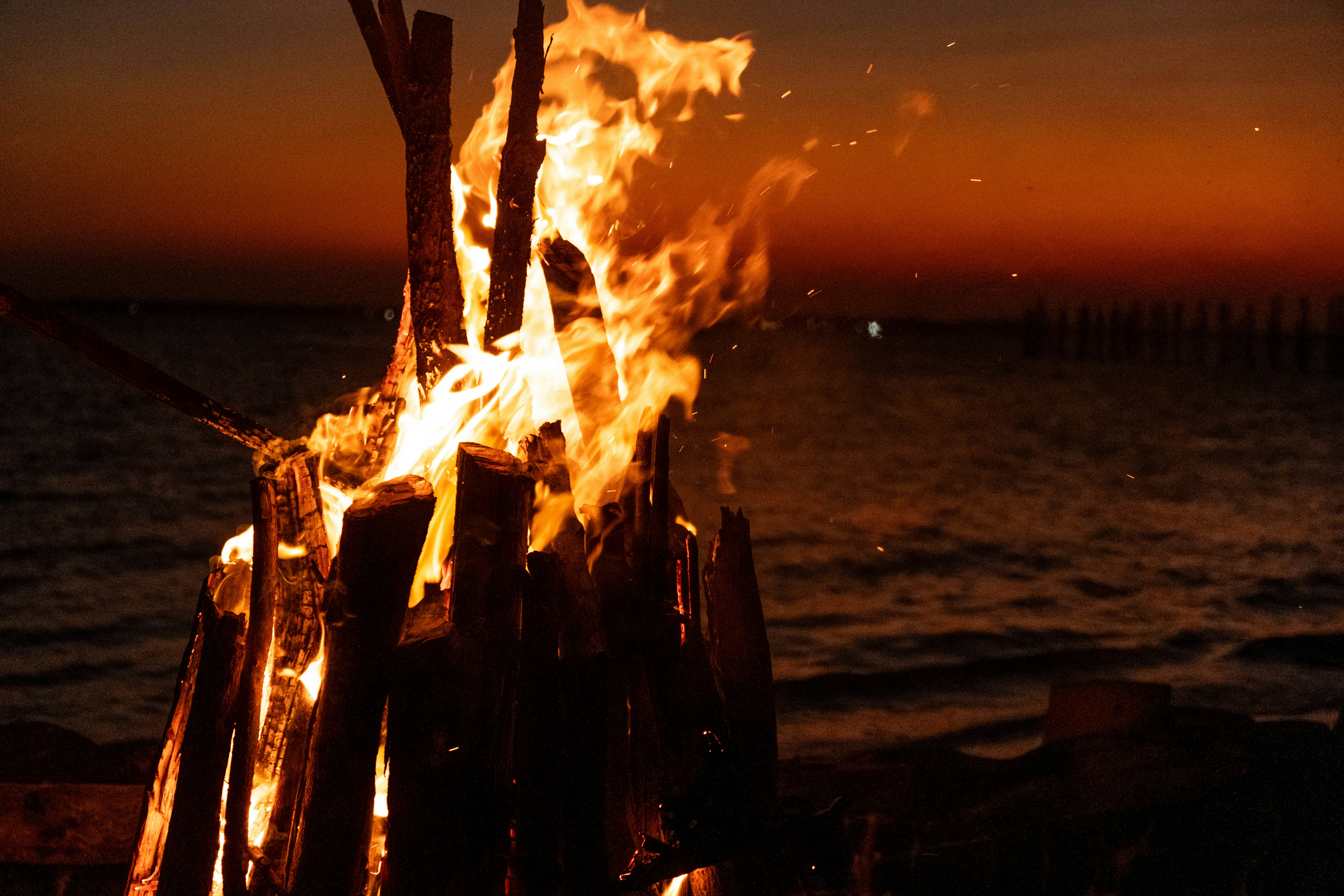 A crackling bonfire along a darkened shoreline at sunset with wooden logs ablaze and subtle distant lights on the horizon.