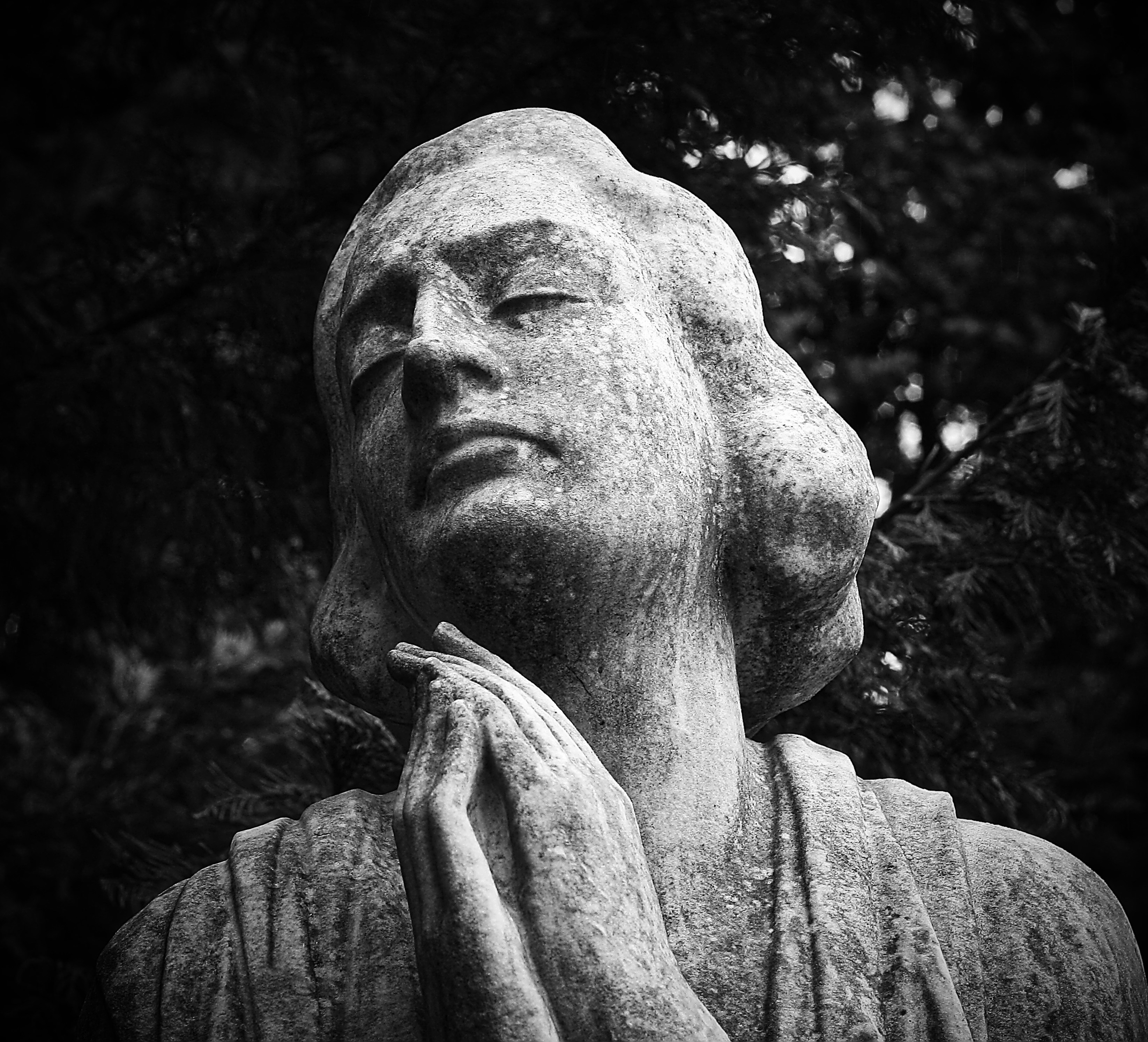Monumental stone statue captured in grayscale, hands clasped in prayer with a tranquil, upward-tilted gaze set against a dark, leafy background.