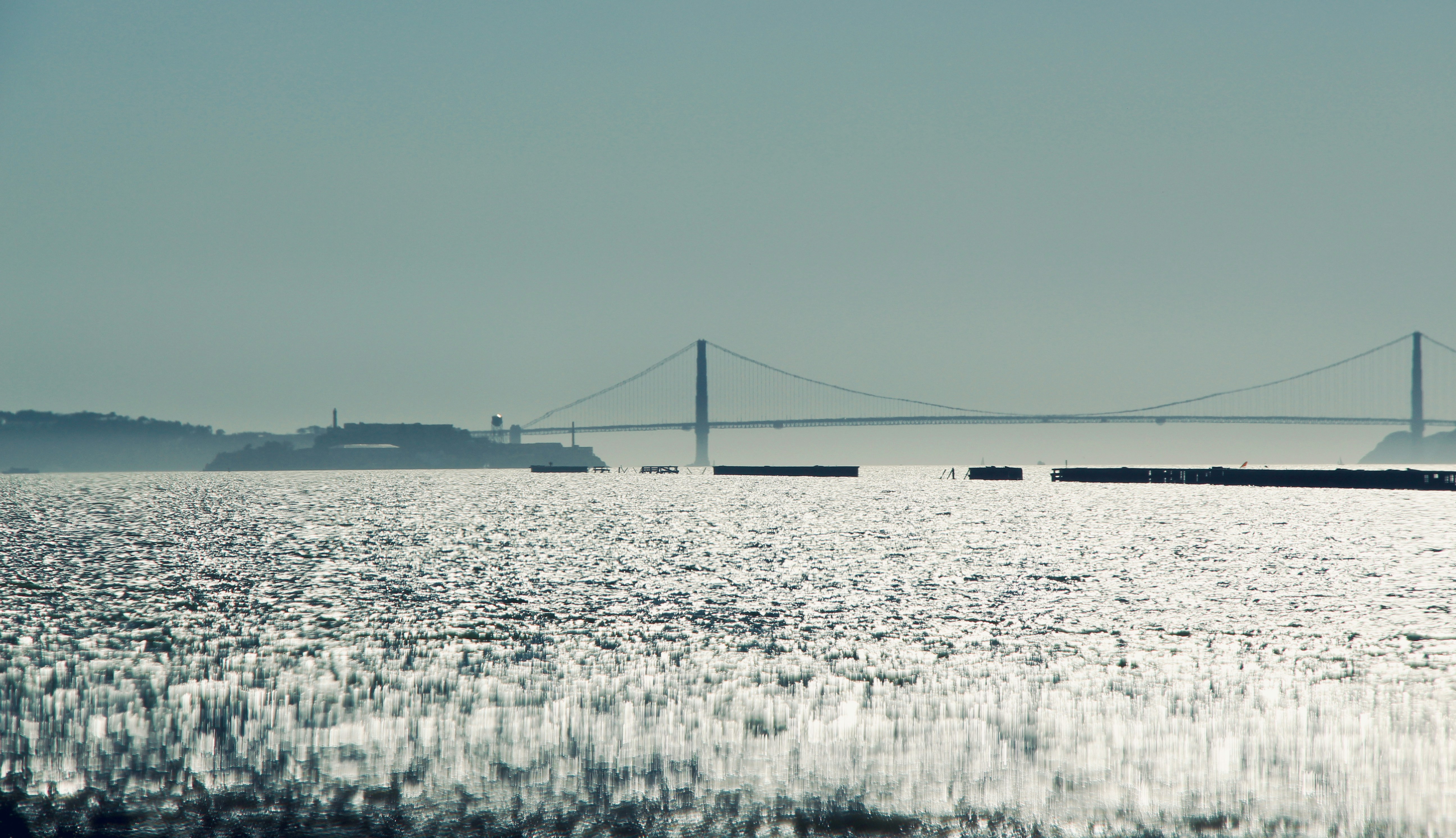 Coastal scene with a distant suspension bridge, shimmering water, and a calm horizon under soft light.