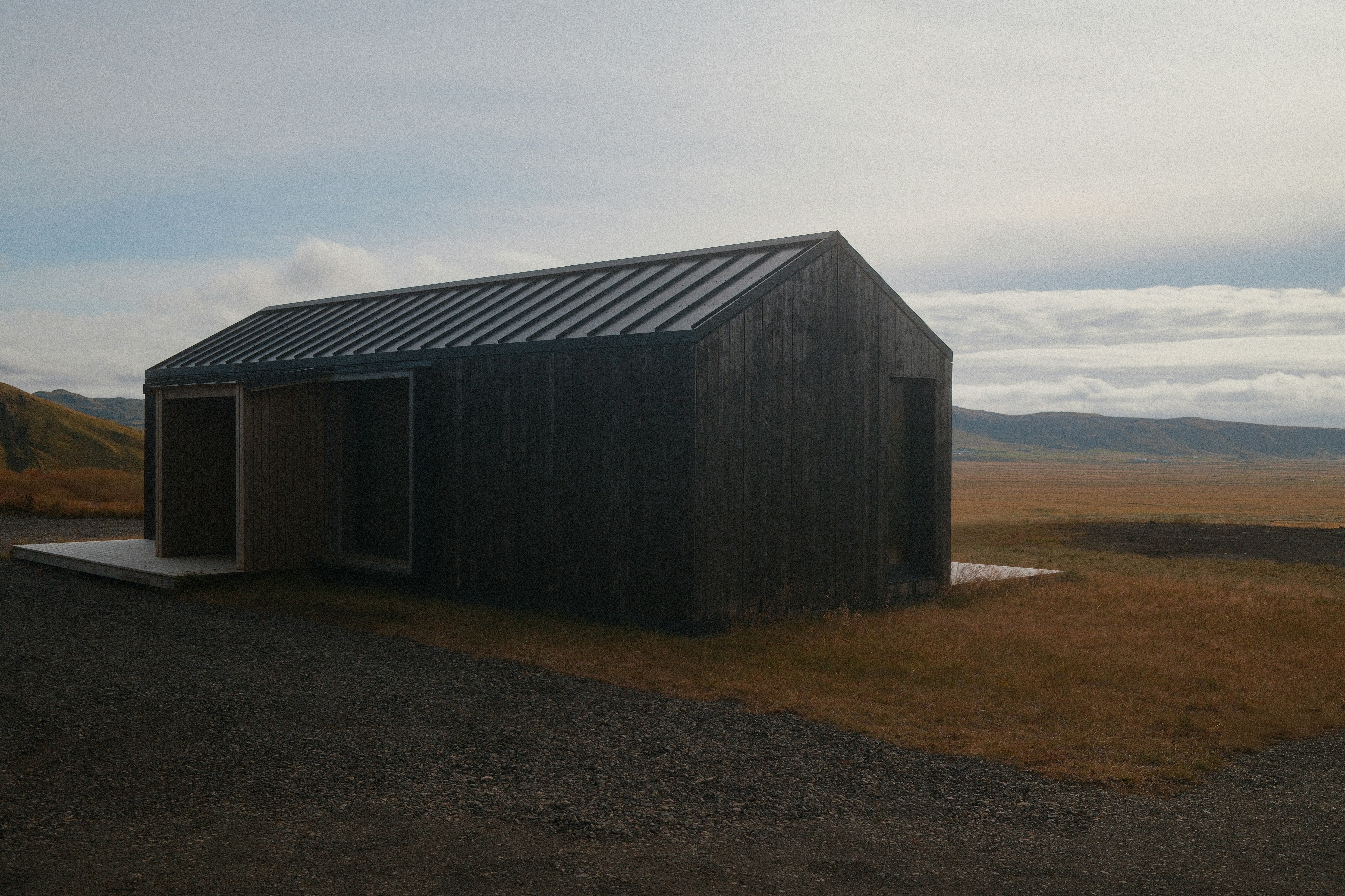 Dark wooden shed sits on moorland with a gravel approach under a muted sky. Rural landscape with distant hills and brown grass, a lone structure giving a sense of solitude.