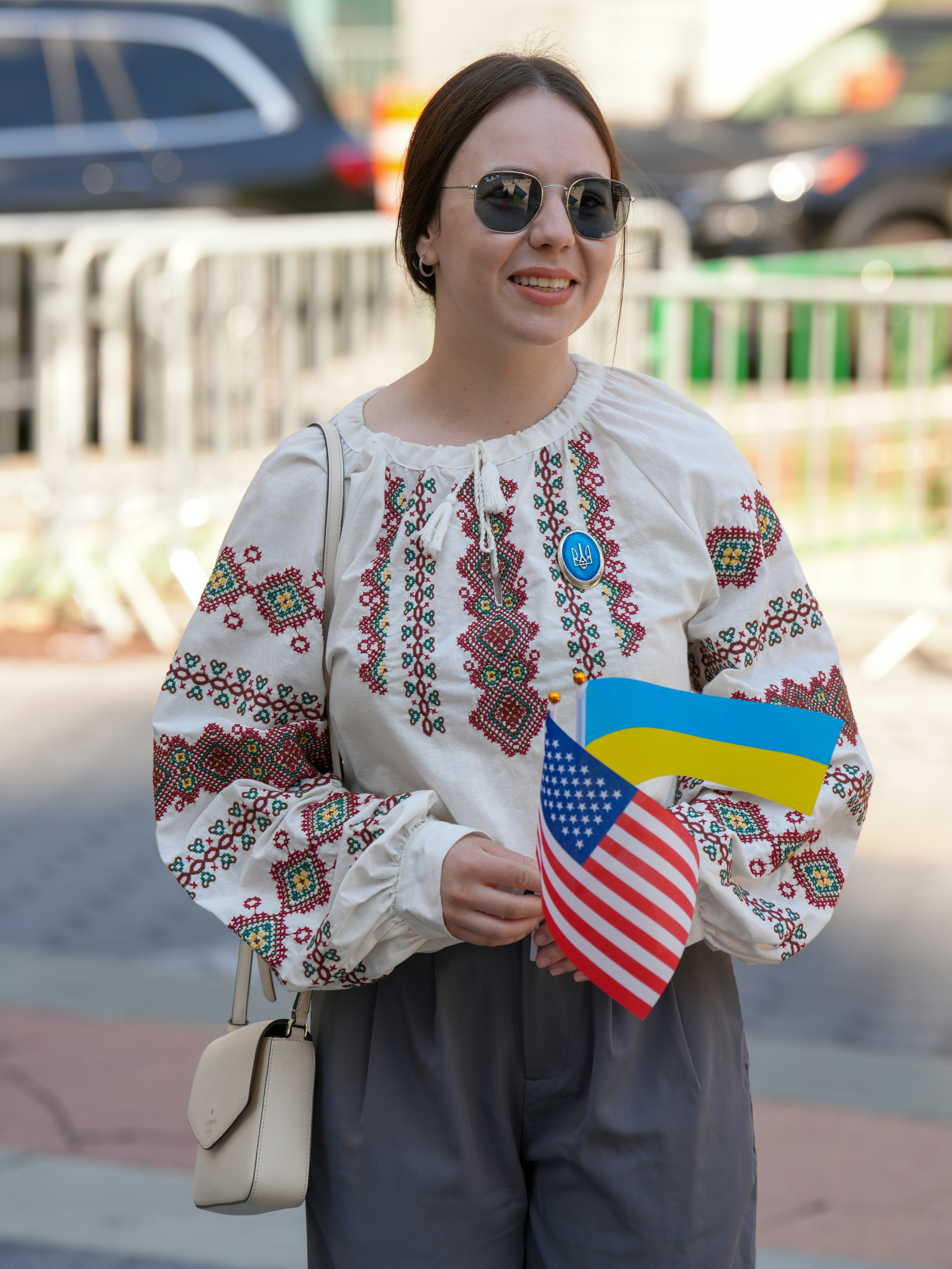A smiling young woman in round sunglasses stands on a city street wearing a traditional Ukrainian embroidered blouse with red and green cross-stitch patterns and a blue trident pin. She holds small Ukrainian and American flags in one hand, with a white shoulder bag at her side and blurred cars and metal barriers behind her. Photo: Olek Buzunov (@buzunov.photoart)