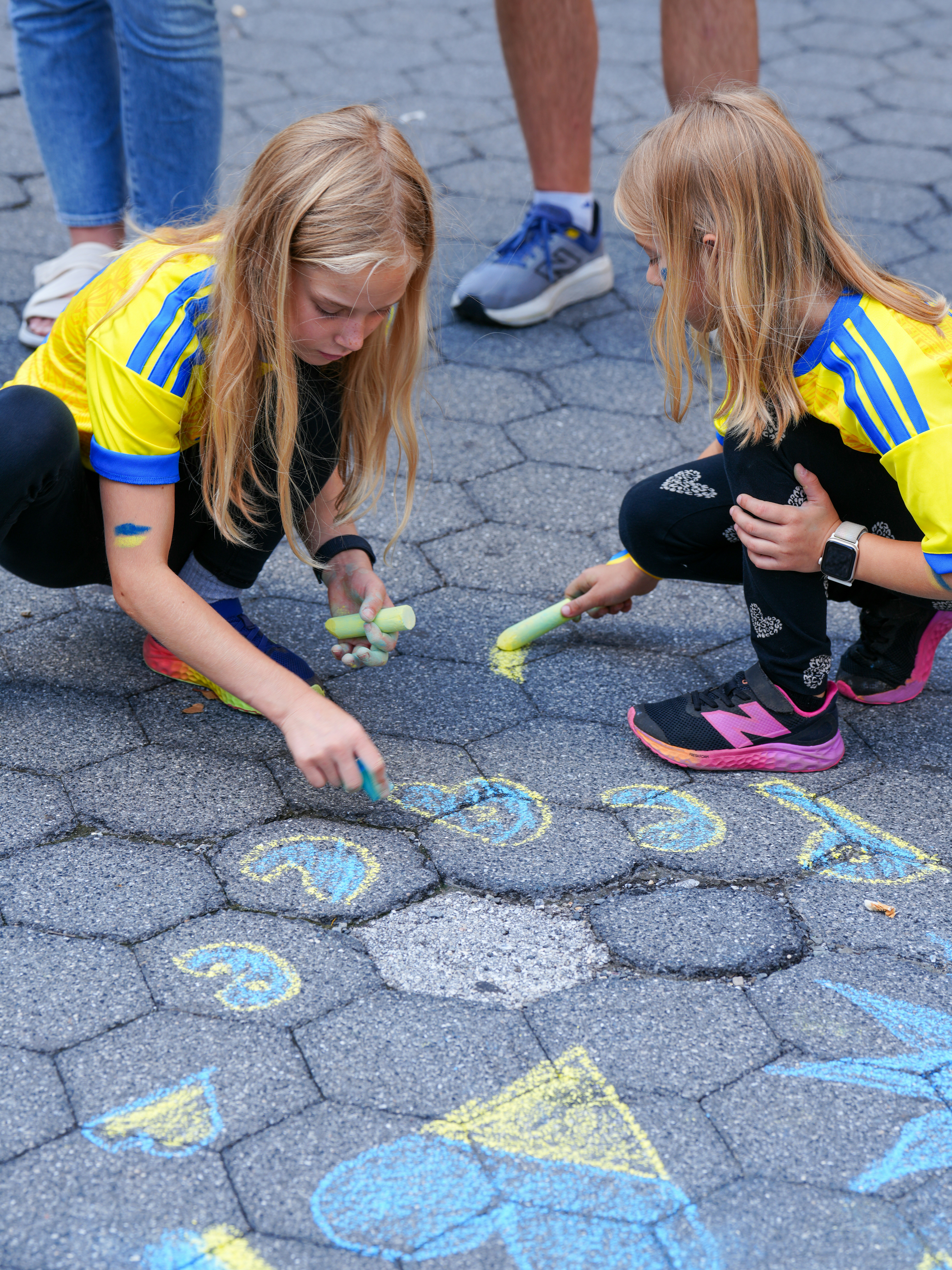 Families gather on a cobblestone city plaza as children draw with chalk on the pavement. A girl in a yellow-and-blue Ukraine soccer jersey kneels to sketch, while adults in matching blue jerseys stand nearby holding small Ukrainian and American flags under scaffolding-lined buildings. Scenes from a Ukrainian anti-war rally in downtown Manhattan, where demonstrators gathered with flags and signs to protest the war and show solidarity with Ukraine.