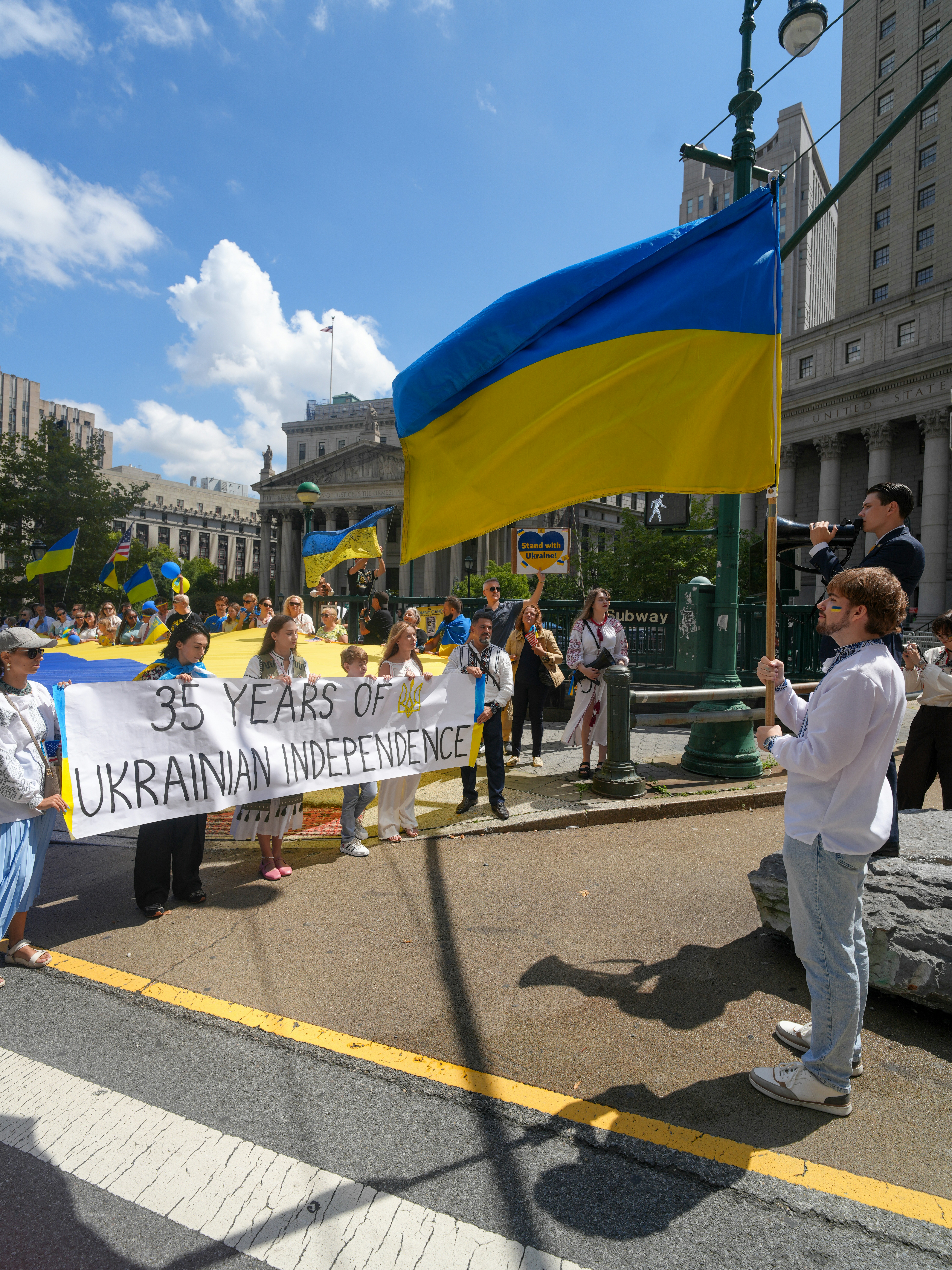 A man with blue-and-yellow face paint waves a large Ukrainian flag beside a subway entrance as marchers hold a "35 Years of Ukrainian Independence" banner across the street. A speaker with a megaphone and supporters carrying a giant flag gather near the columned courthouse in Foley Square on a bright, cloud-streaked day. Photo: Olek Buzunov (@buzunov.photoart)