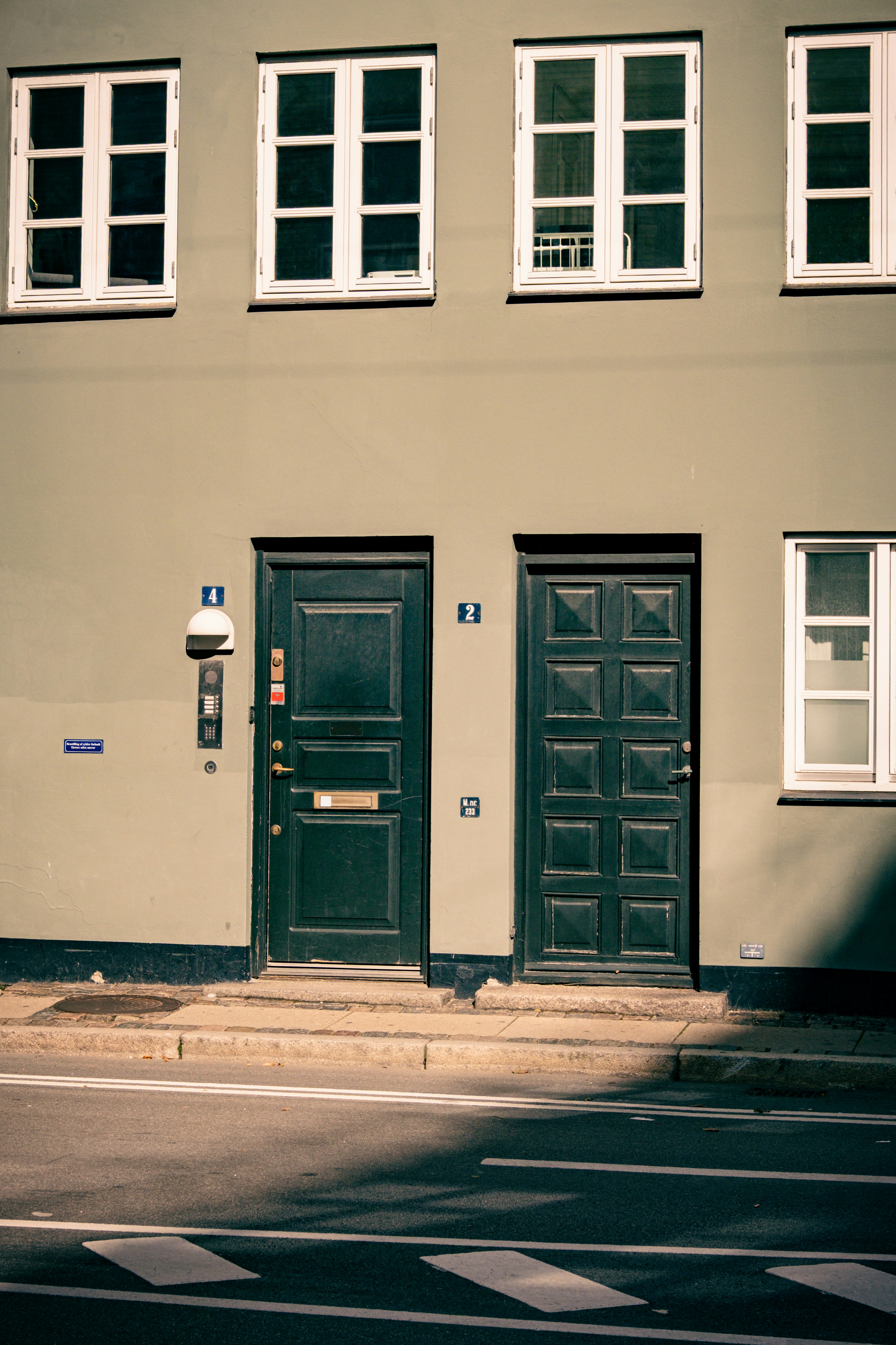 Two dark doors sit under a pale facade with windows above, casting a long street shadow and a subtle diagonal of light on the sidewalk.