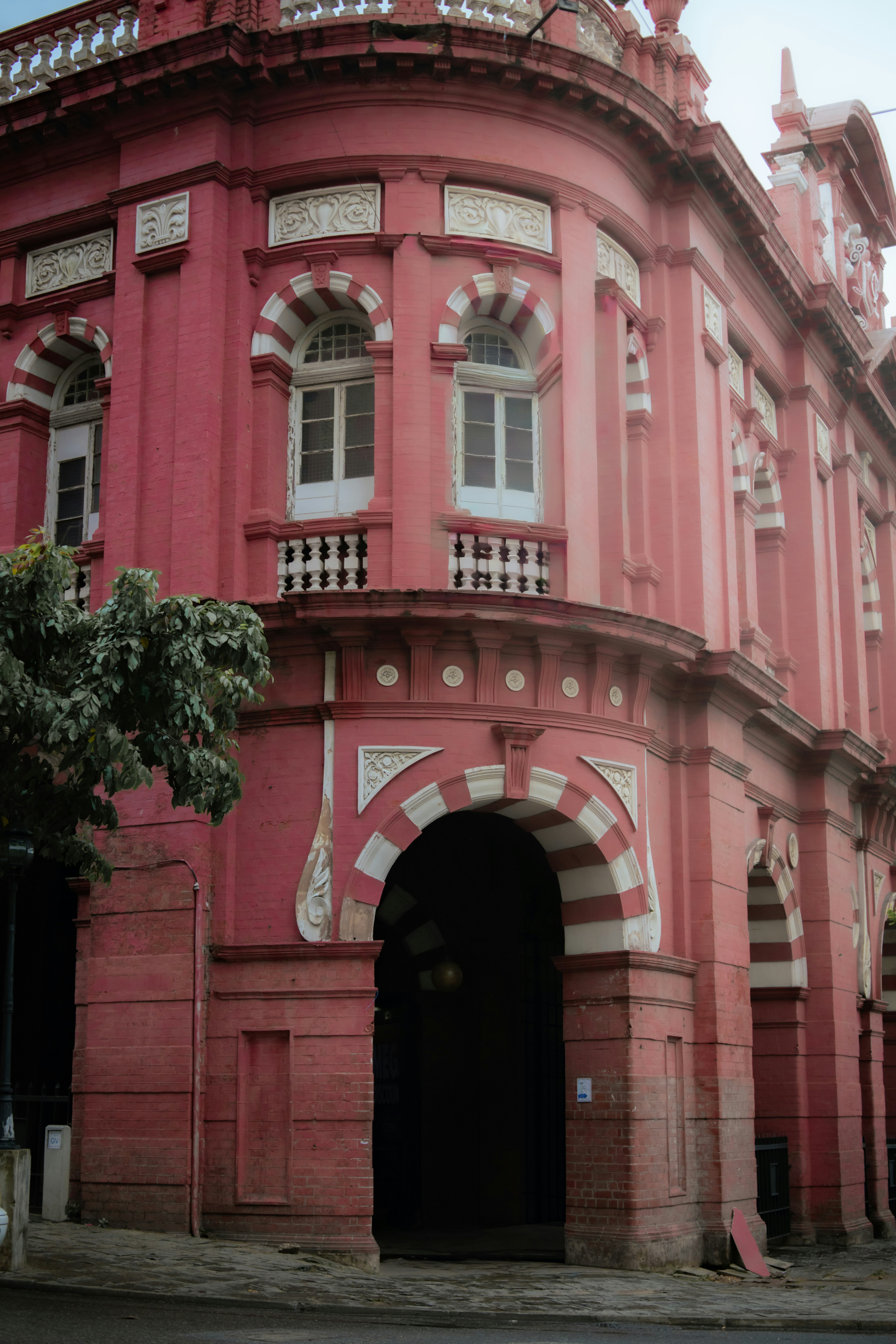 A grand red-brick facade with striped arches and arched windows dominates the frame; a dark arched entrance anchors the lower portion, while a tree adds natural contrast to the historic structure.