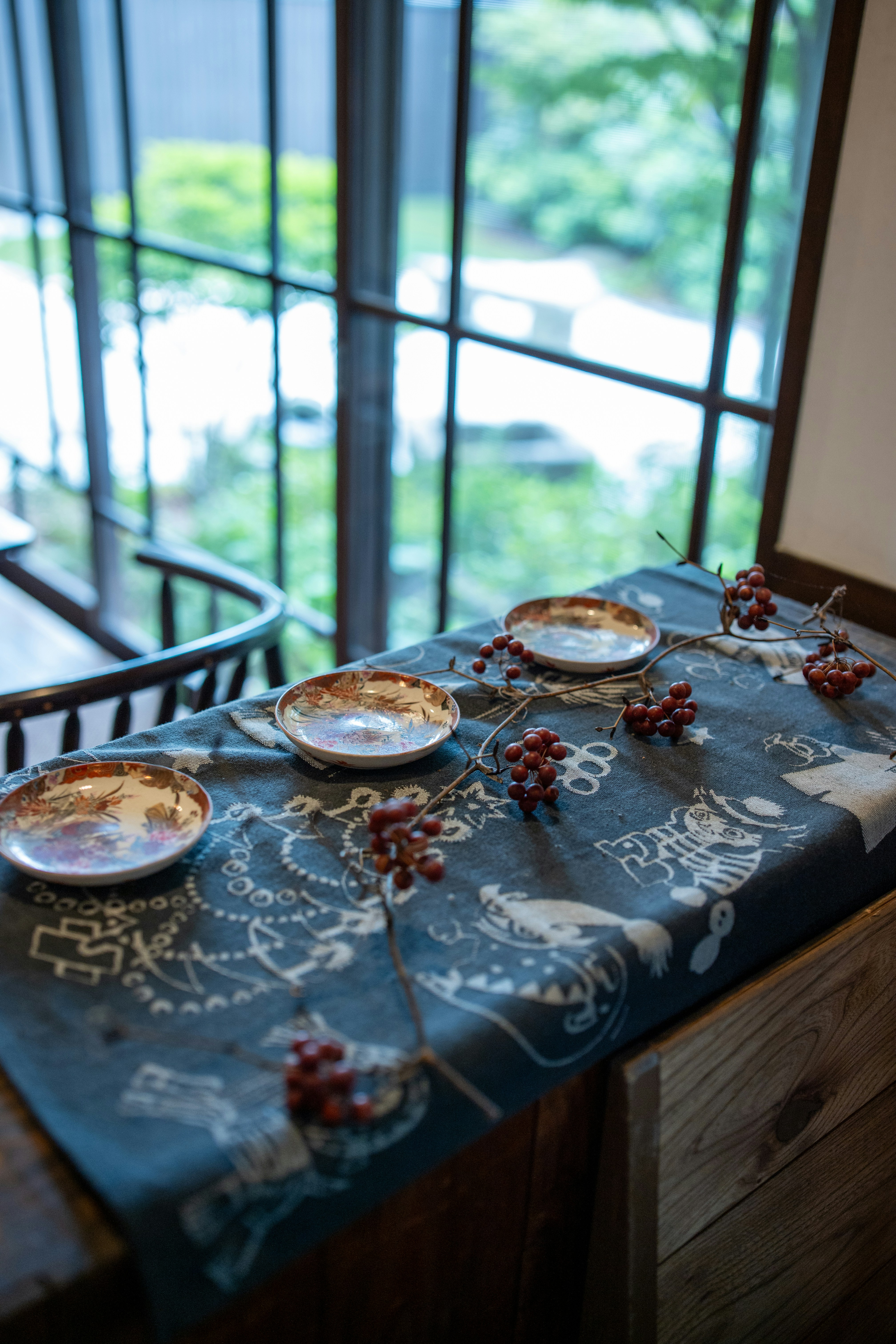 Blue patterned tablecloth with small ceramic dishes and red berries is set along a wooden railing, near a large window; soft natural light highlights the tabletop arrangement. This is a photograph.