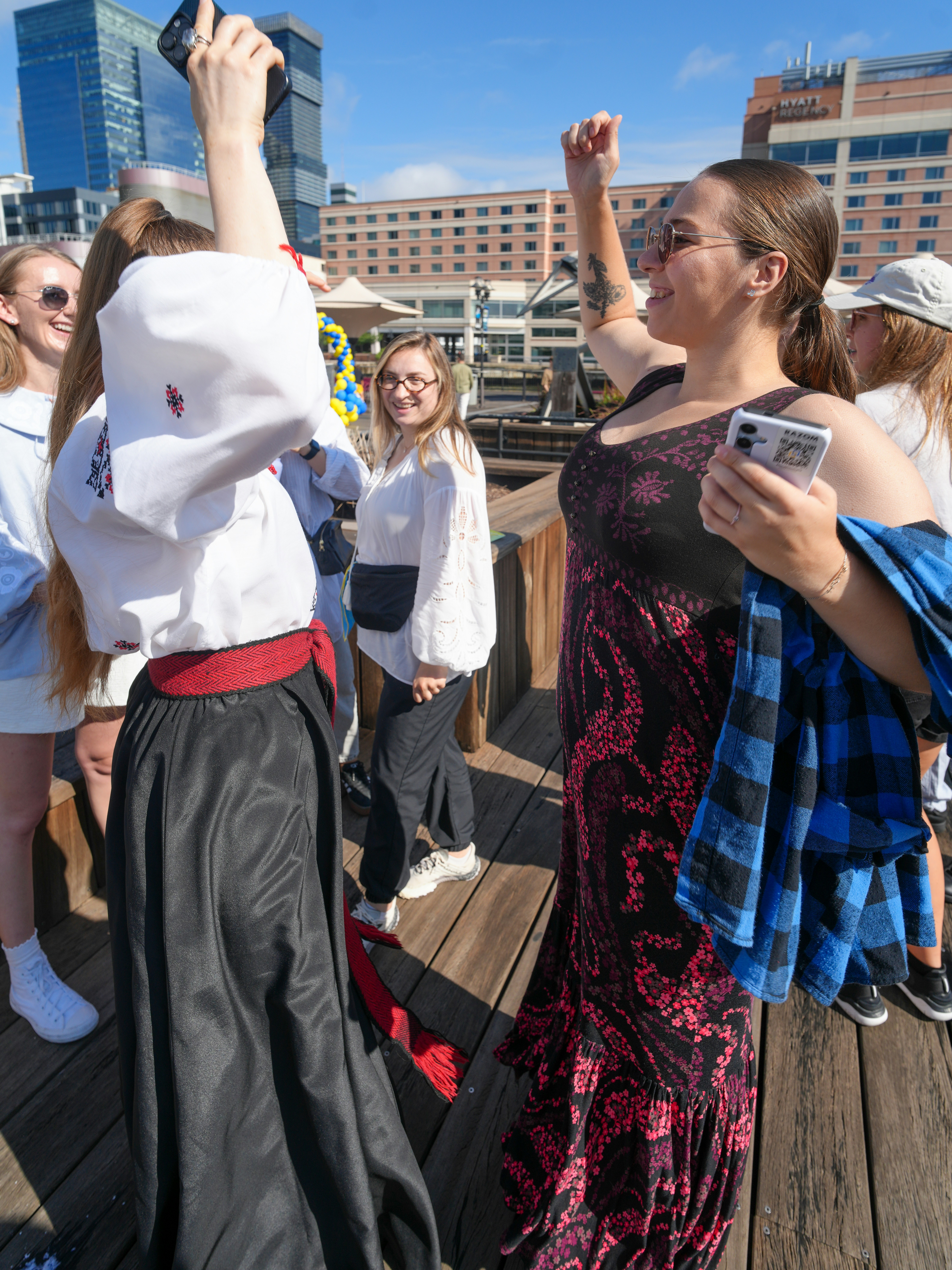 Women dance joyfully with raised arms on a sunny waterfront boardwalk, one in a traditional white embroidered blouse and black skirt with a red woven sash, another in a floral maxi dress holding a phone, with modern glass towers and hotel buildings behind them. Scenes from the 2026 Ukrainian Charity Vyshyvanka Run along the Exchange Place promenade in Jersey City, where runners in traditional embroidered shirts gathered to support Ukraine. Photo: Olek Buzunov (@buzunov.photoart)