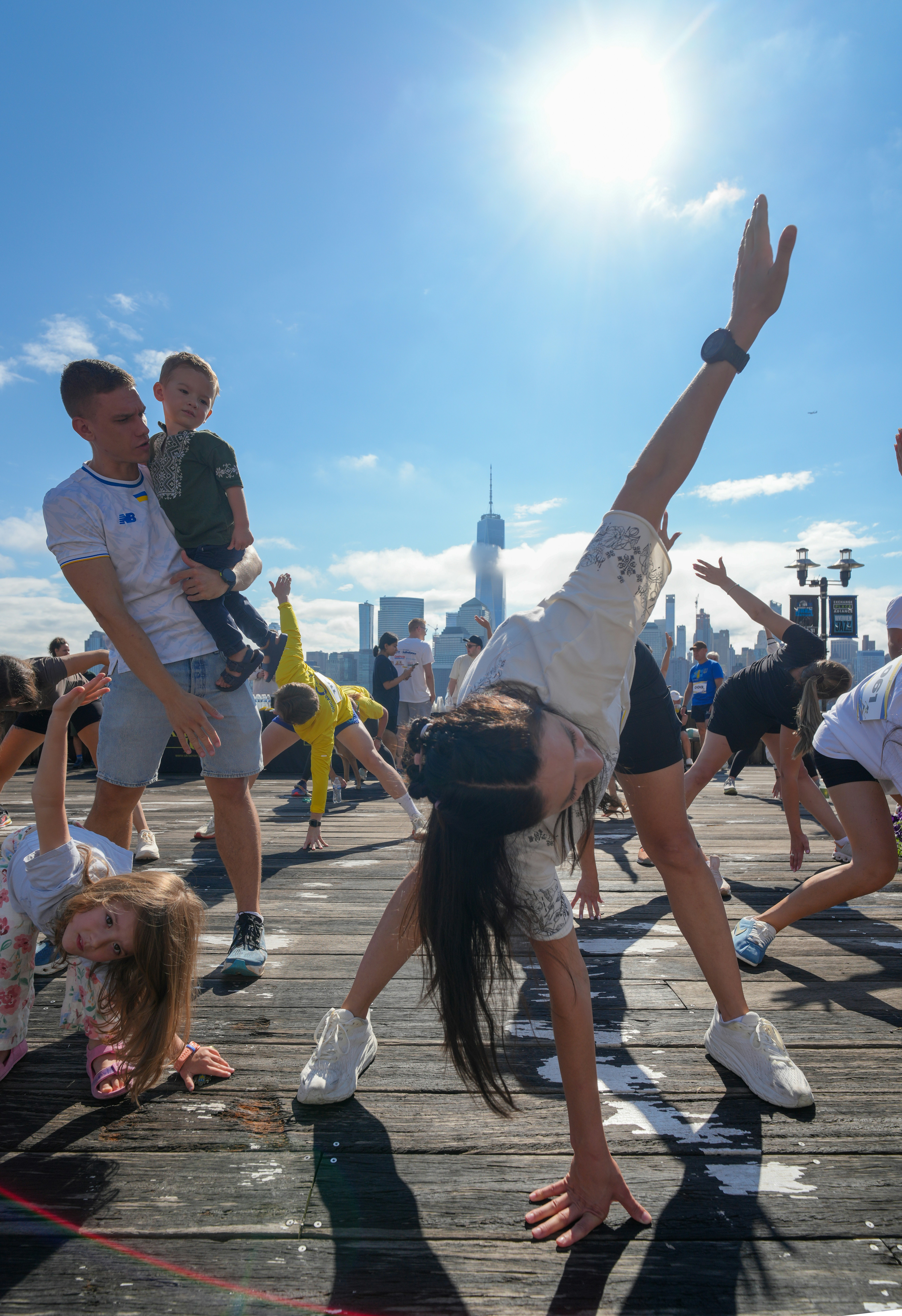Runners warm up in the morning sun on a wooden waterfront promenade, with the Lower Manhattan skyline and One World Trade Center rising across the river. A woman stretches into a deep standing pose while a father holds his toddler and children play nearby at the Ukrainian charity Vyshyvanka Run in Jersey City. Scenes from the 2026 Ukrainian Charity Vyshyvanka Run along the Exchange Place promenade in Jersey City, where runners in traditional embroidered shirts gathered to support Ukraine. Photo: Olek Buzunov (@buzunov.photoart)