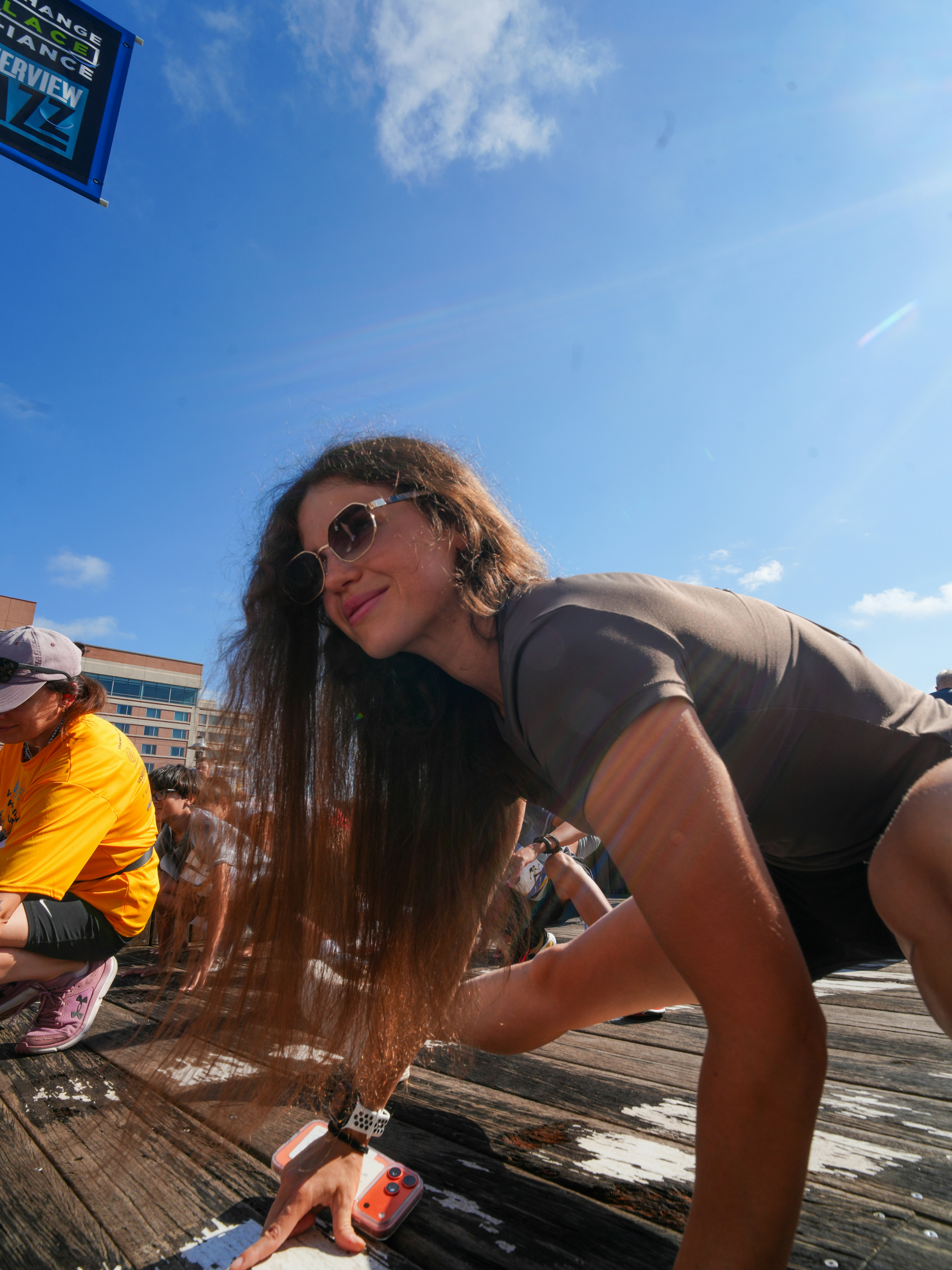 A young woman with long flowing hair and sunglasses smiles while holding a low sprinter's stretch on a weathered wooden promenade at the Ukrainian charity run in Jersey City. Behind her, other participants warm up in the bright morning sunshine. Scenes from the 2026 Ukrainian Charity Vyshyvanka Run along the Exchange Place promenade in Jersey City, where runners in traditional embroidered shirts gathered to support Ukraine. Photo: Olek Buzunov (@buzunov.photoart)