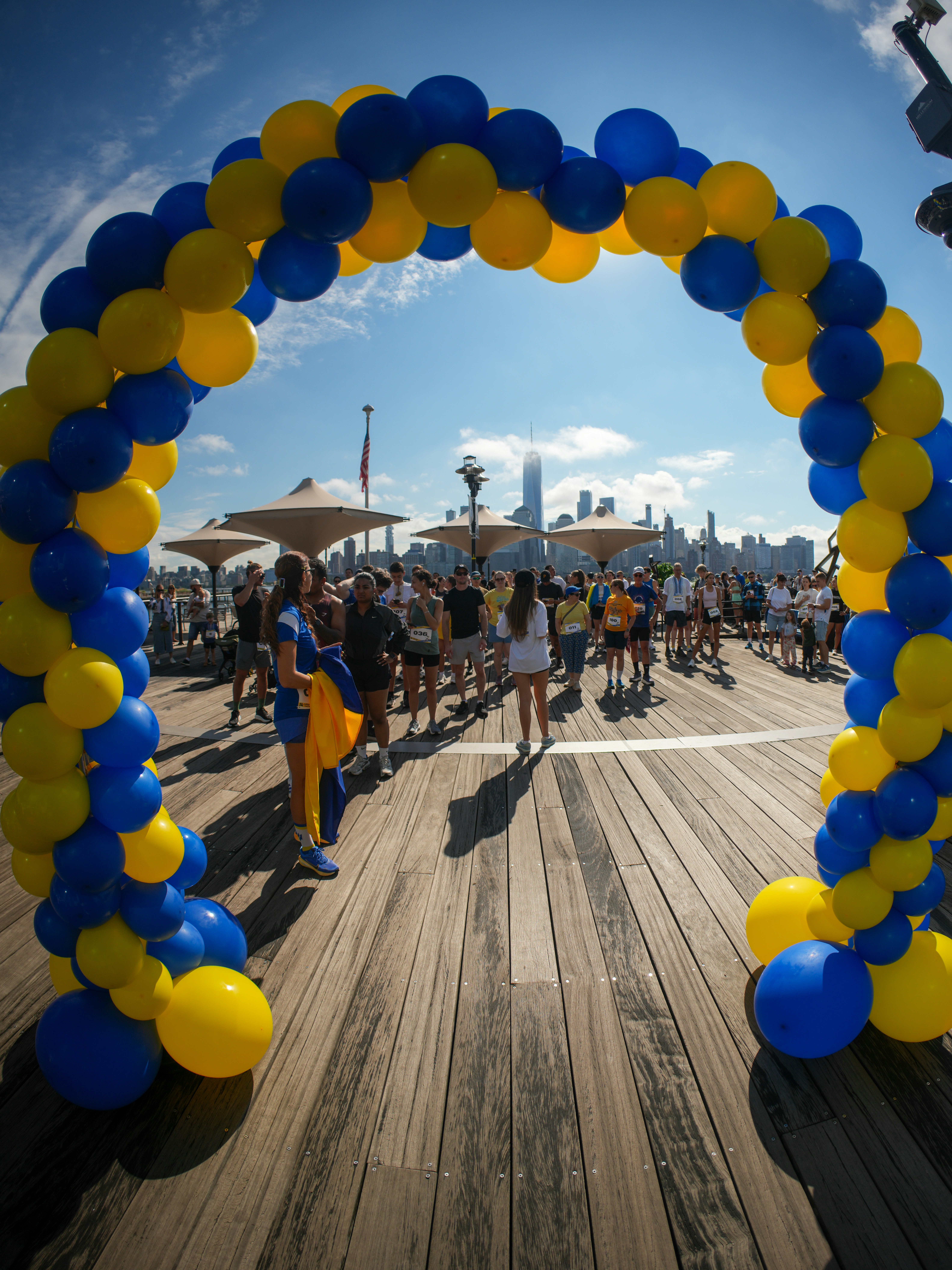 A vertical fisheye view through a towering blue and yellow balloon arch on a wooden pier promenade, where runners with race bibs gather before the Ukrainian Charity Vyshyvanka Run. Beige canopy umbrellas, an American flag, and the sunlit Manhattan skyline with One World Trade Center rise in the background. Scenes from the 2026 Ukrainian Charity Vyshyvanka Run along the Exchange Place promenade in Jersey City, where runners in traditional embroidered shirts gathered to support Ukraine. Photo: Olek Buzunov (@buzunov.photoart)