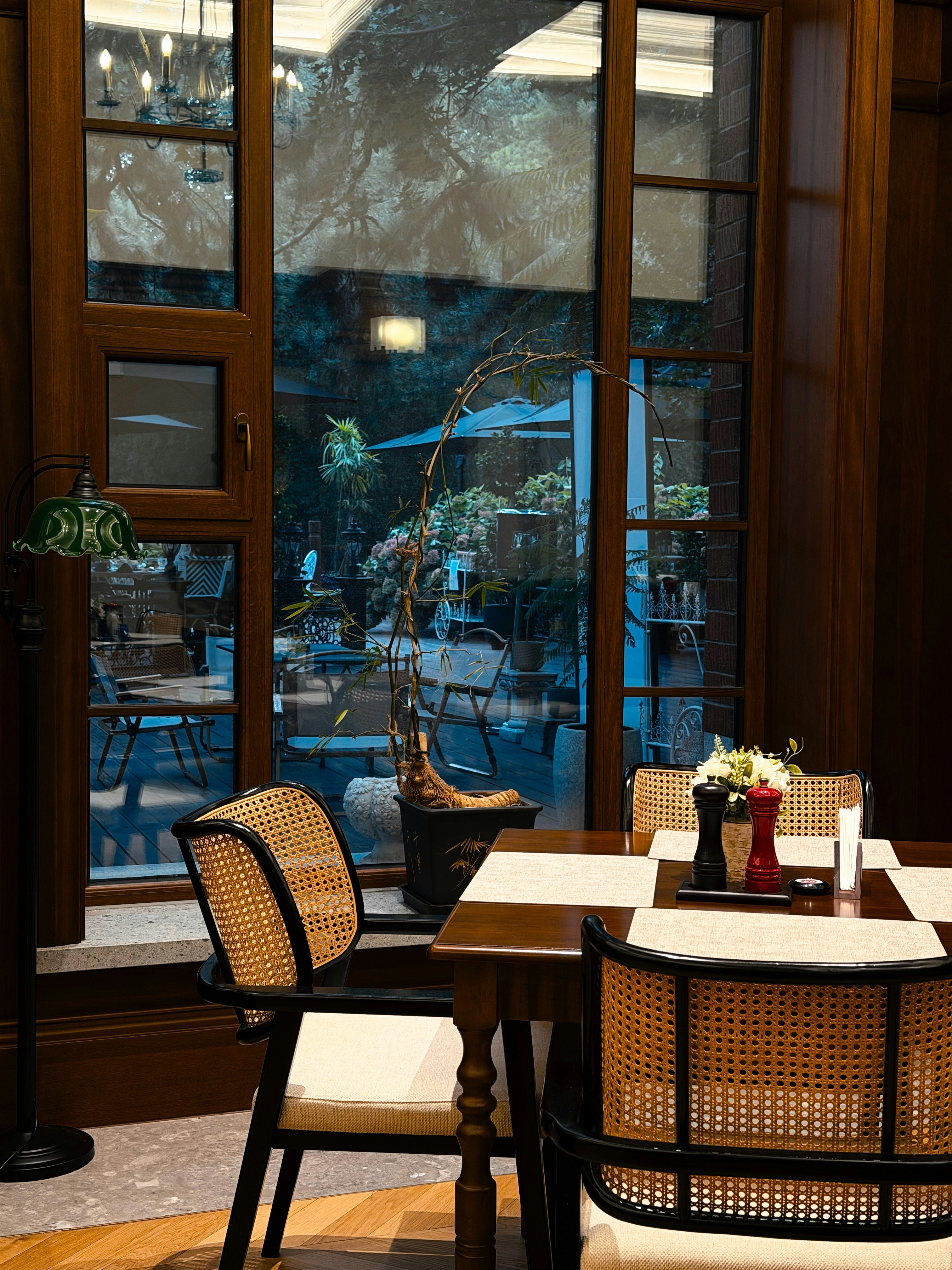 Warm-lit bistro seating by large windows; a wooden table with caned chairs and a small centerpiece hints at a calm dining space.