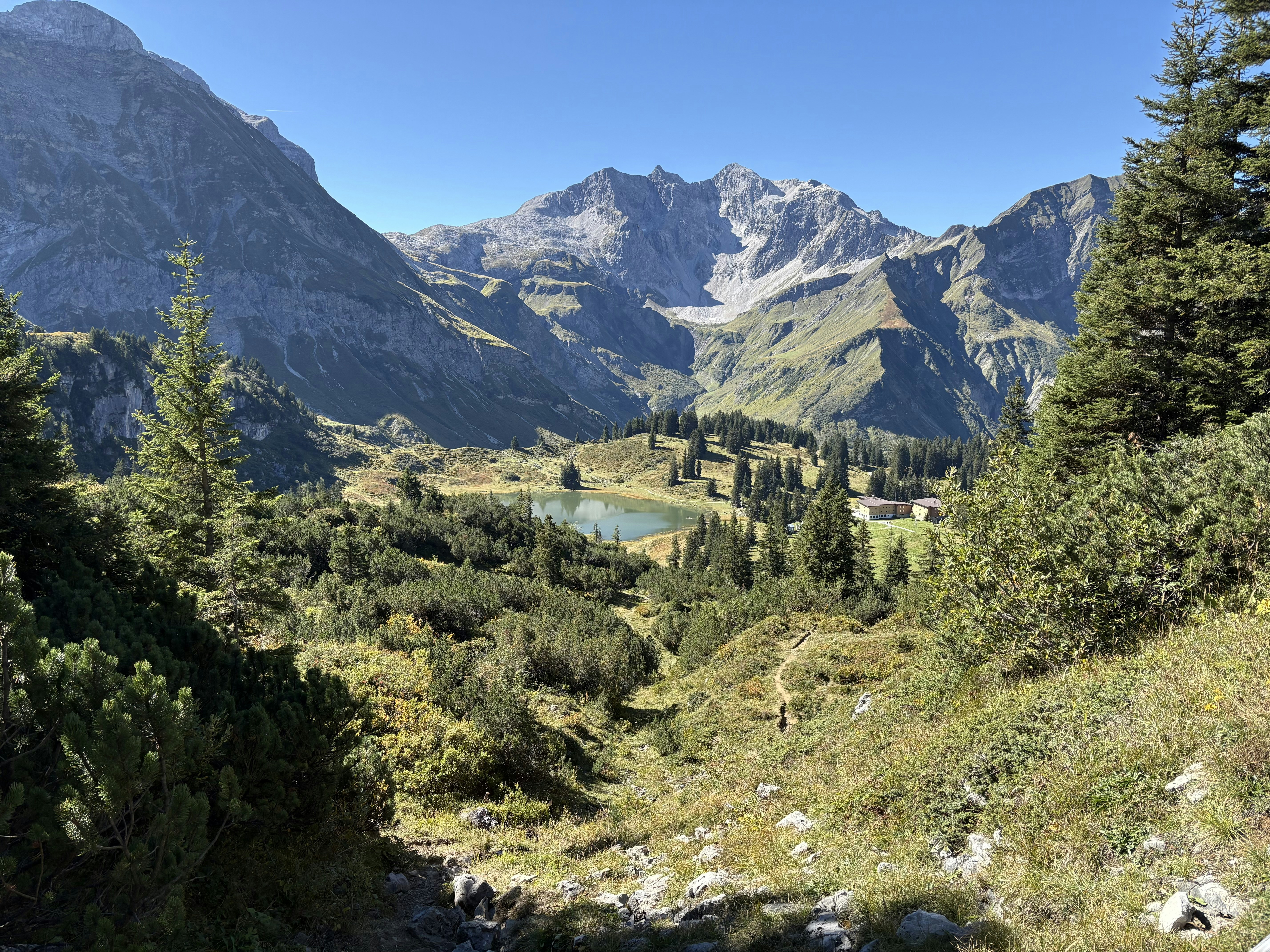 Snow-dusted mountain peaks frame a sunlit alpine valley with a small pond and winding trail; a serene landscape scene highlighting peaks, meadow, and conifers.