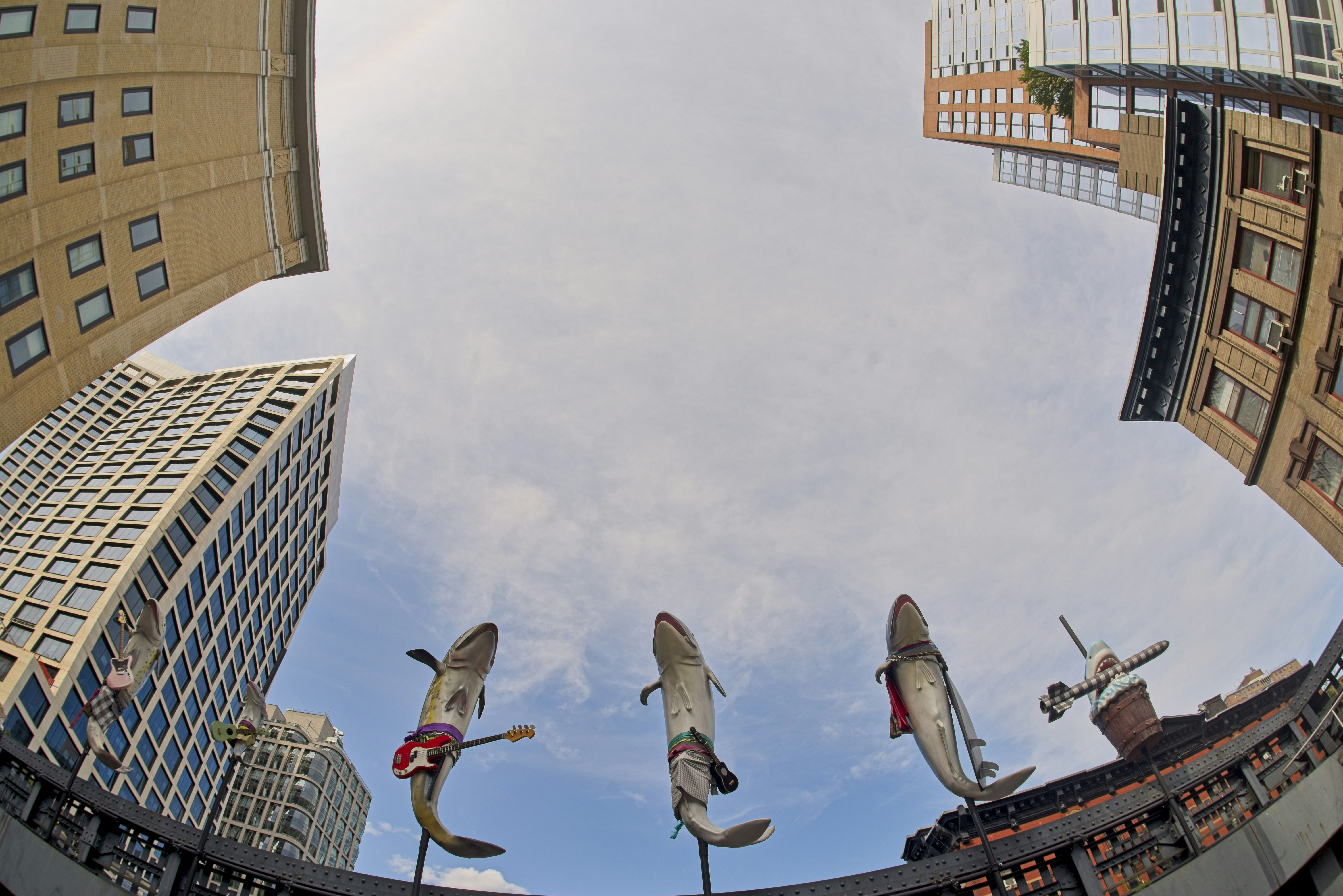 High Line Observation Deck on a warm summer day in July. Looking straight up through a fisheye lens, playful shark sculptures rise on tall poles above the steel railings of this elevated park in Manhattan. Dressed with scarves and bandanas, the sharks carry colorful guitars as if forming a quirky rock band in the sky. A whimsical ice cream cone pierced by a rocket adds to the fun. Surrounding towers of brick, stone and glass curve inward around a bright, cloud-streaked sky, creating a lively, surreal urban scene.